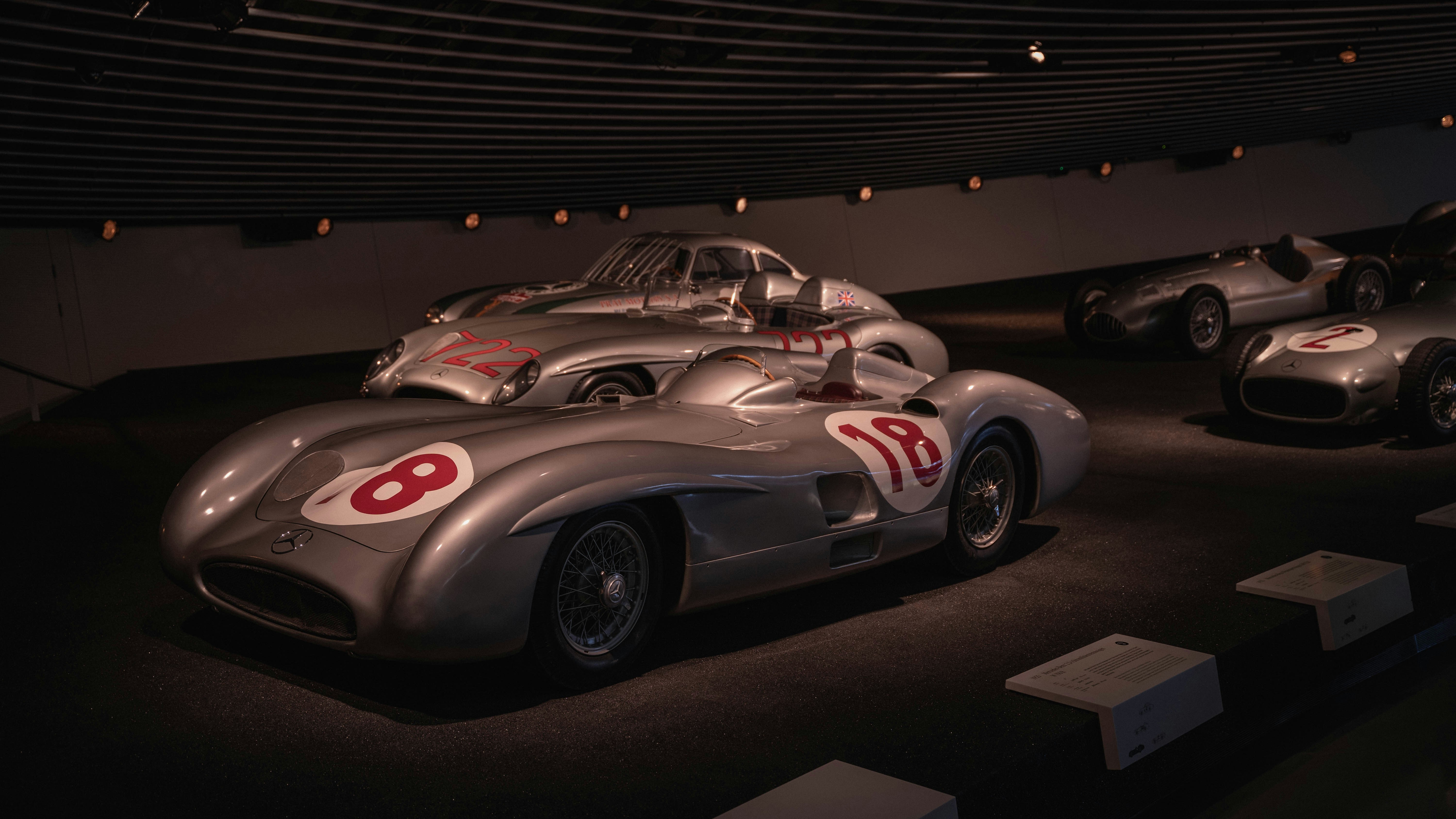 Vintage silver racing cars displayed in a dimly lit exhibition hall.
