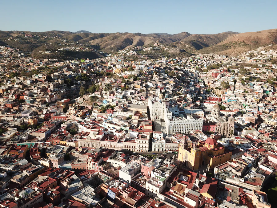 Wide aerial view of a sprawling city packed into a valley, with a prominent cathedral and rolling hills in the background. Sunlight casts clear shadows across the colorful rooftops.