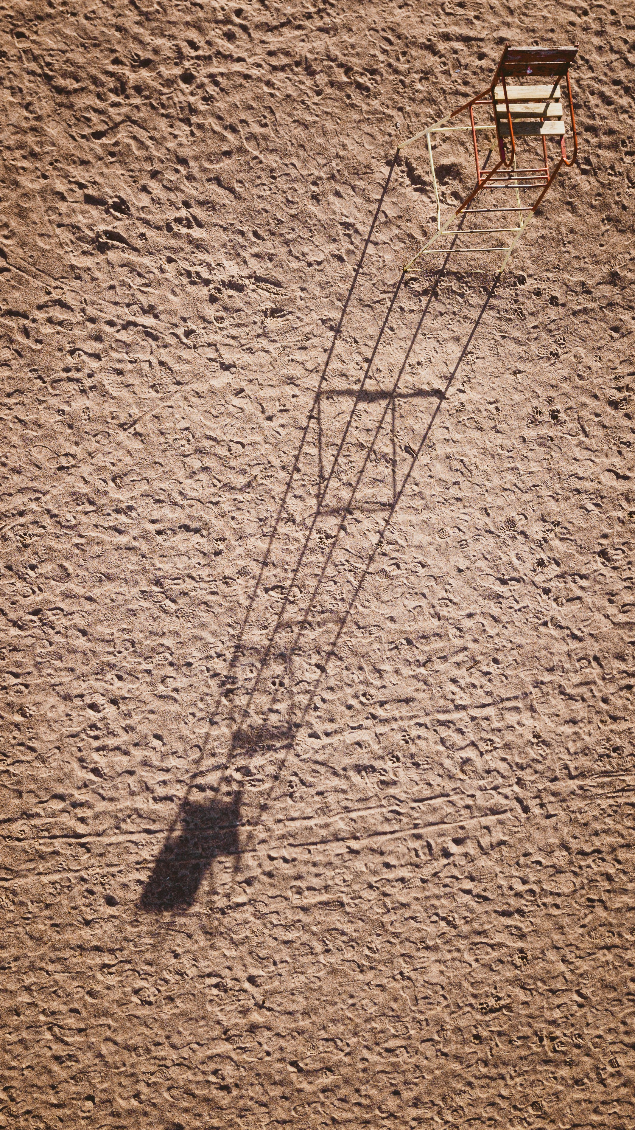 A lifeguard chair casts a long shadow on the sandy beach, highlighting the texture of the sand beneath. 