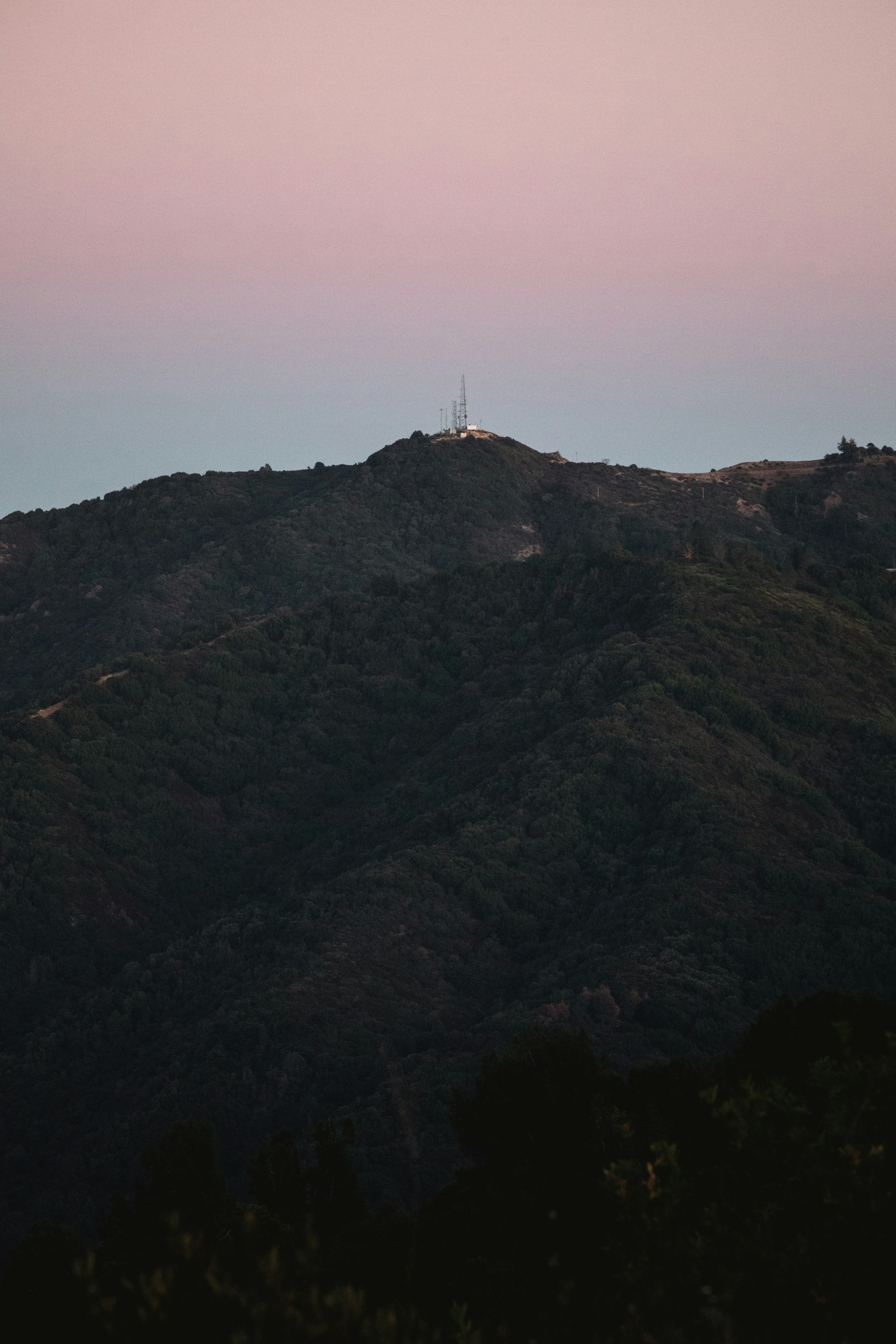 A distant mountain peak crowned with a communication tower, bathed in soft twilight hues. The lush terrain below contrasts with the serene sky above.