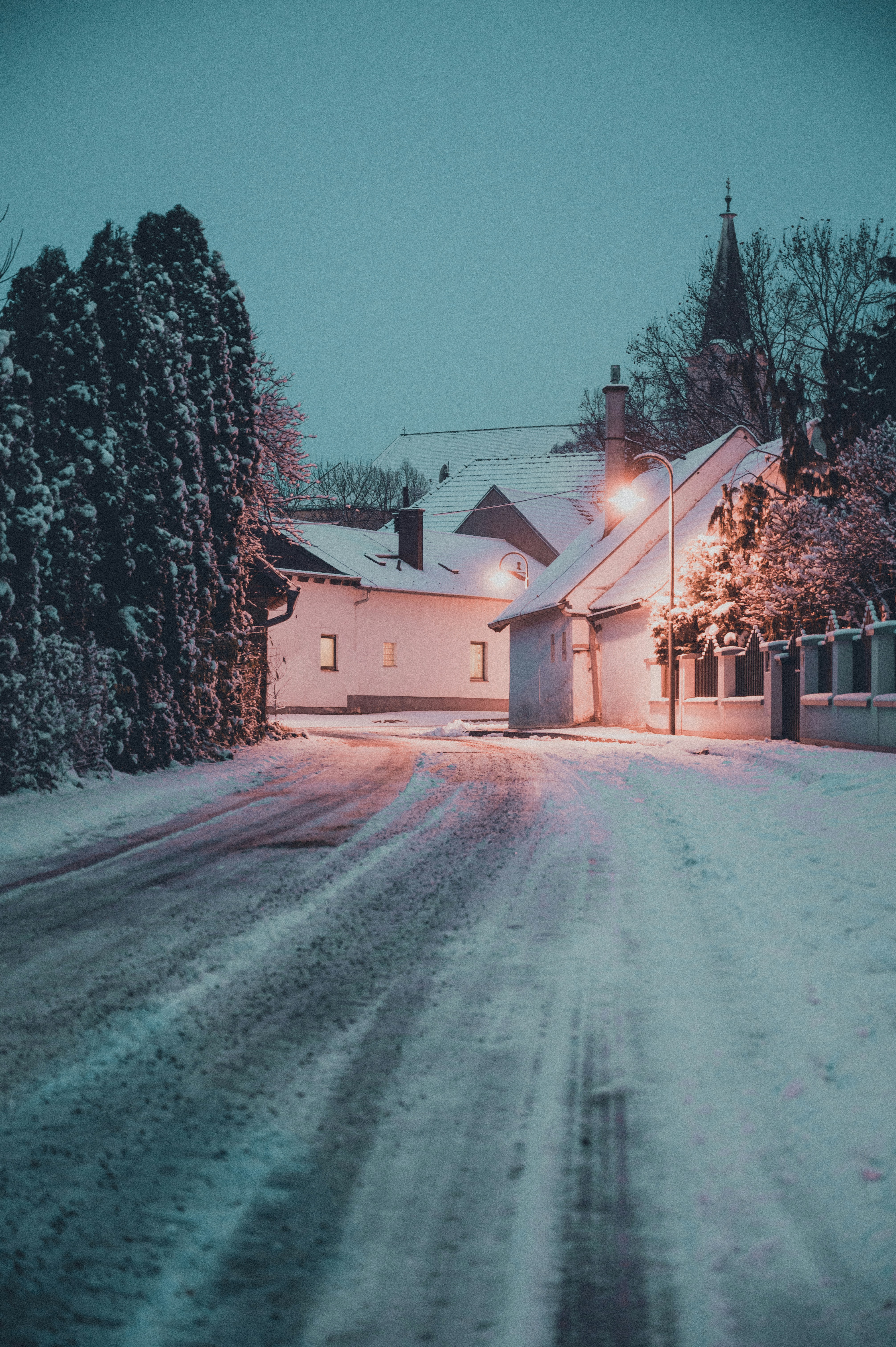Snow-covered street illuminated by soft streetlight, flanked by frosted trees and quaint houses in a serene winter landscape.