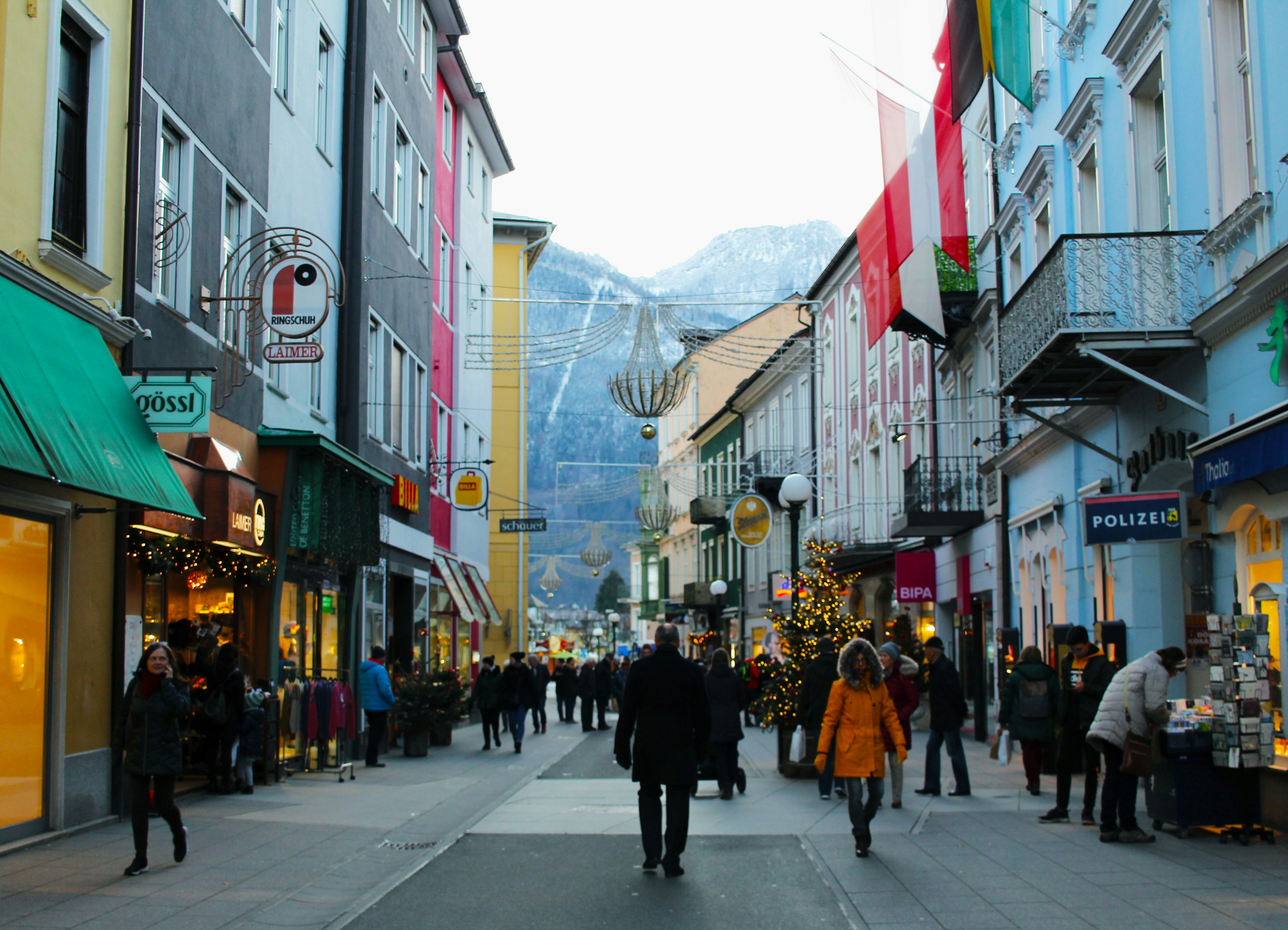 Busy pedestrian street flanked by colorful buildings with snow-capped mountains in the background.