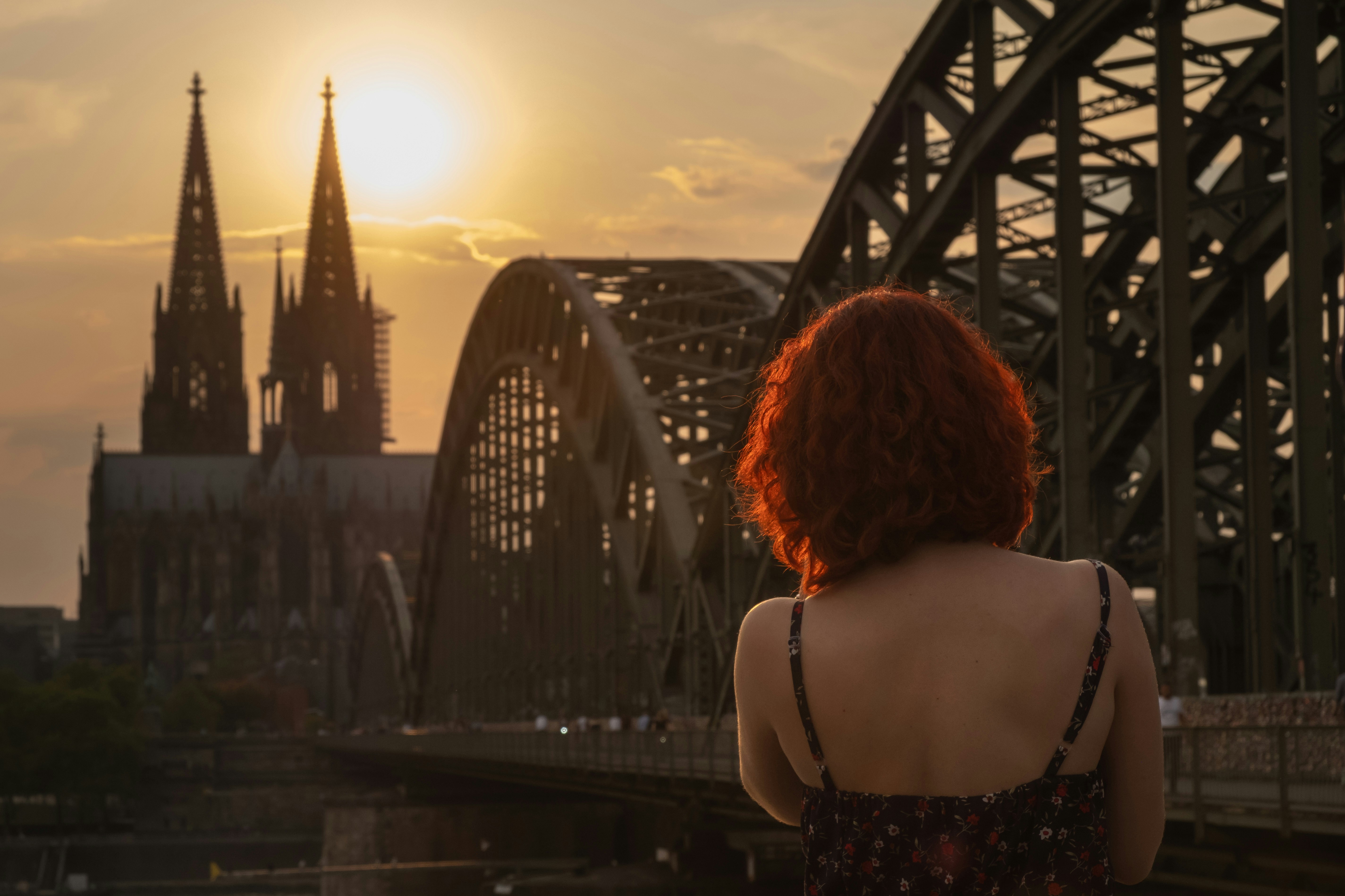 Woman with red hair gazing at a cathedral and bridge under a setting sun.