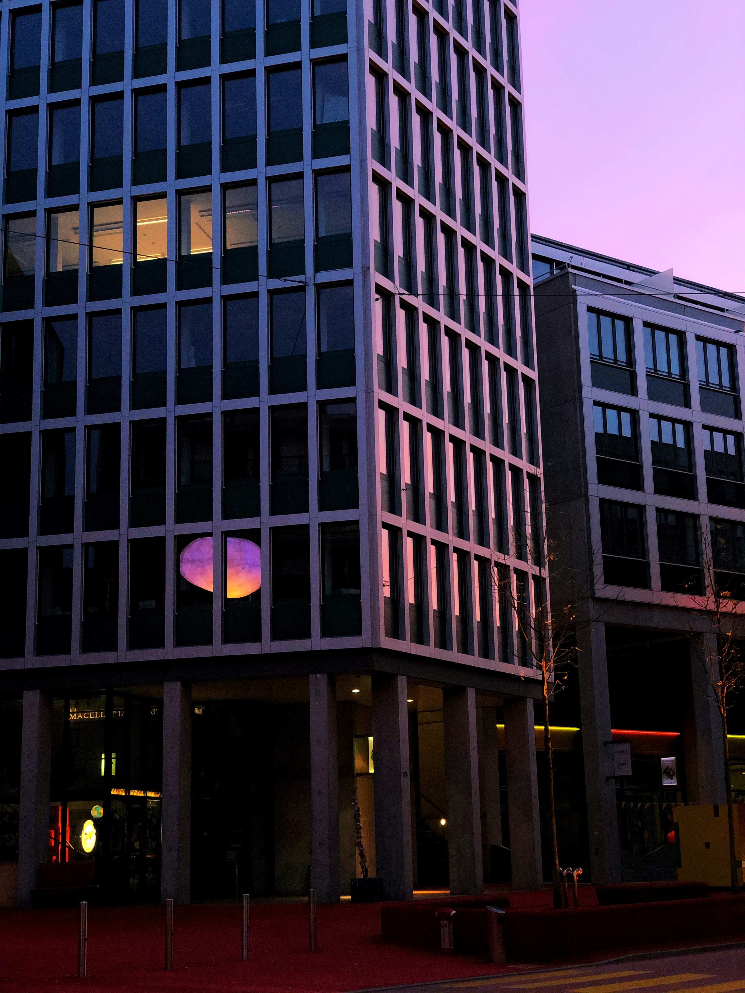 A modern building with a striking reflection of the moon captured in its glass facade during twilight.