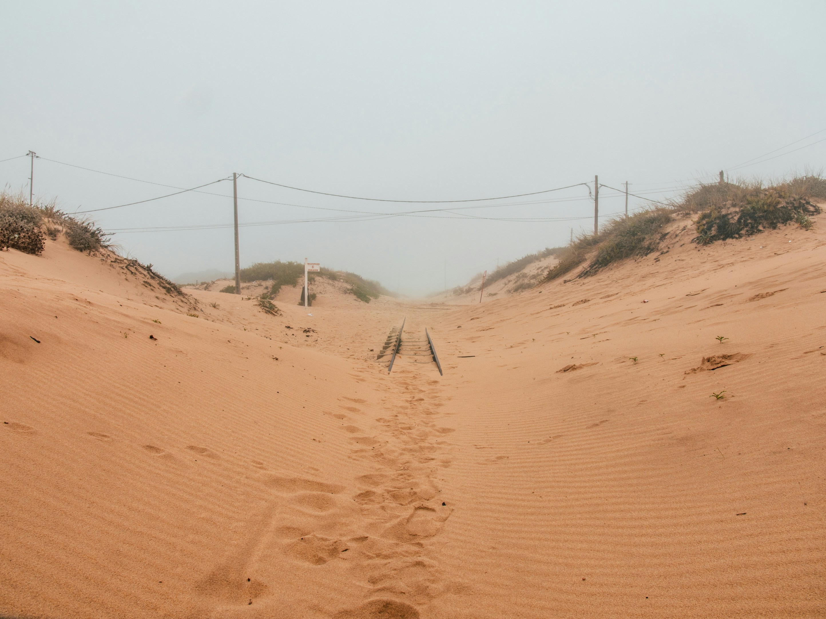 Footprints tracing a path through sandy dunes under a foggy sky, with distant power lines fading into the haze.
