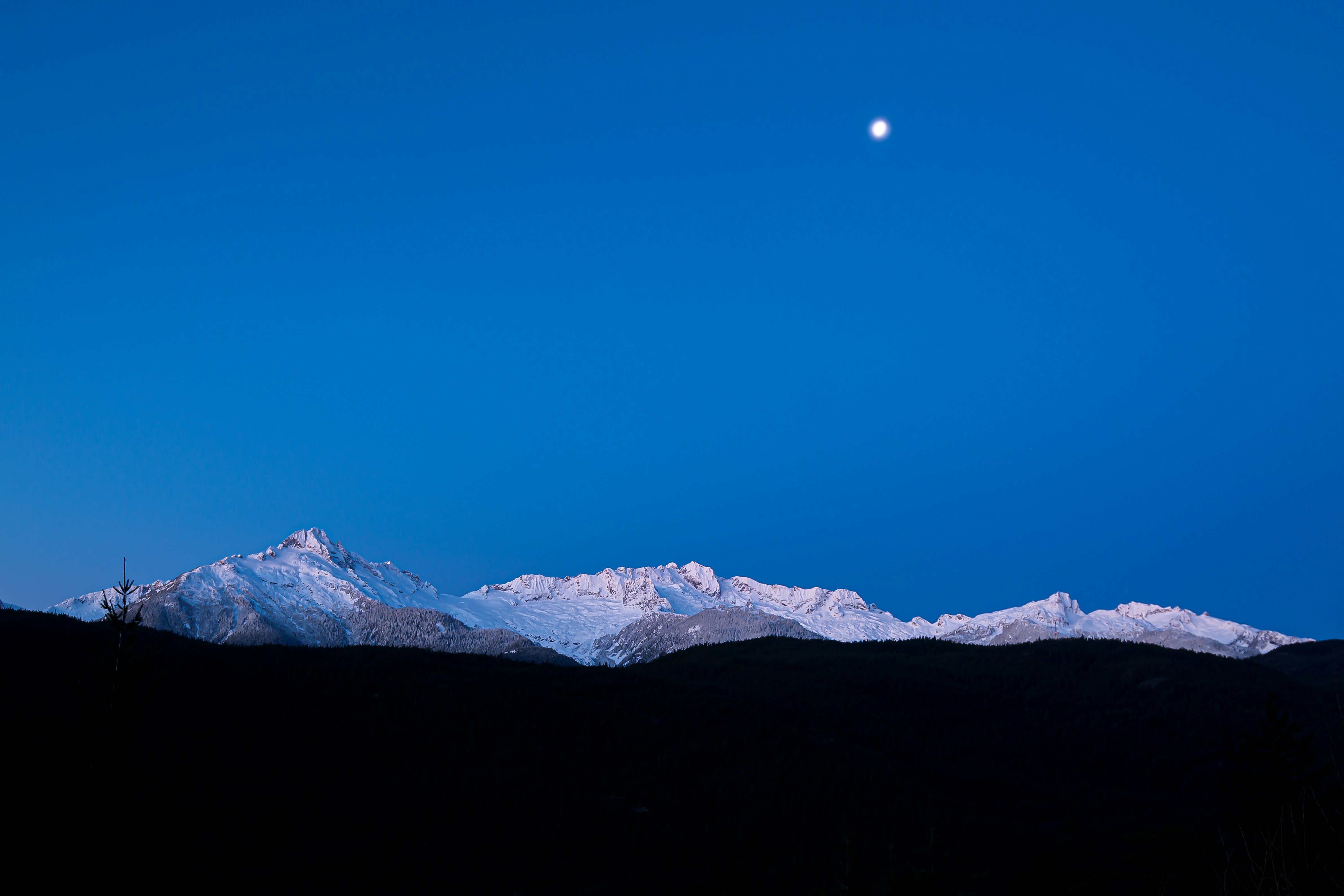 Snow-capped mountains under a clear blue sky with a bright moon above. The scene captures the tranquility of nature at twilight.