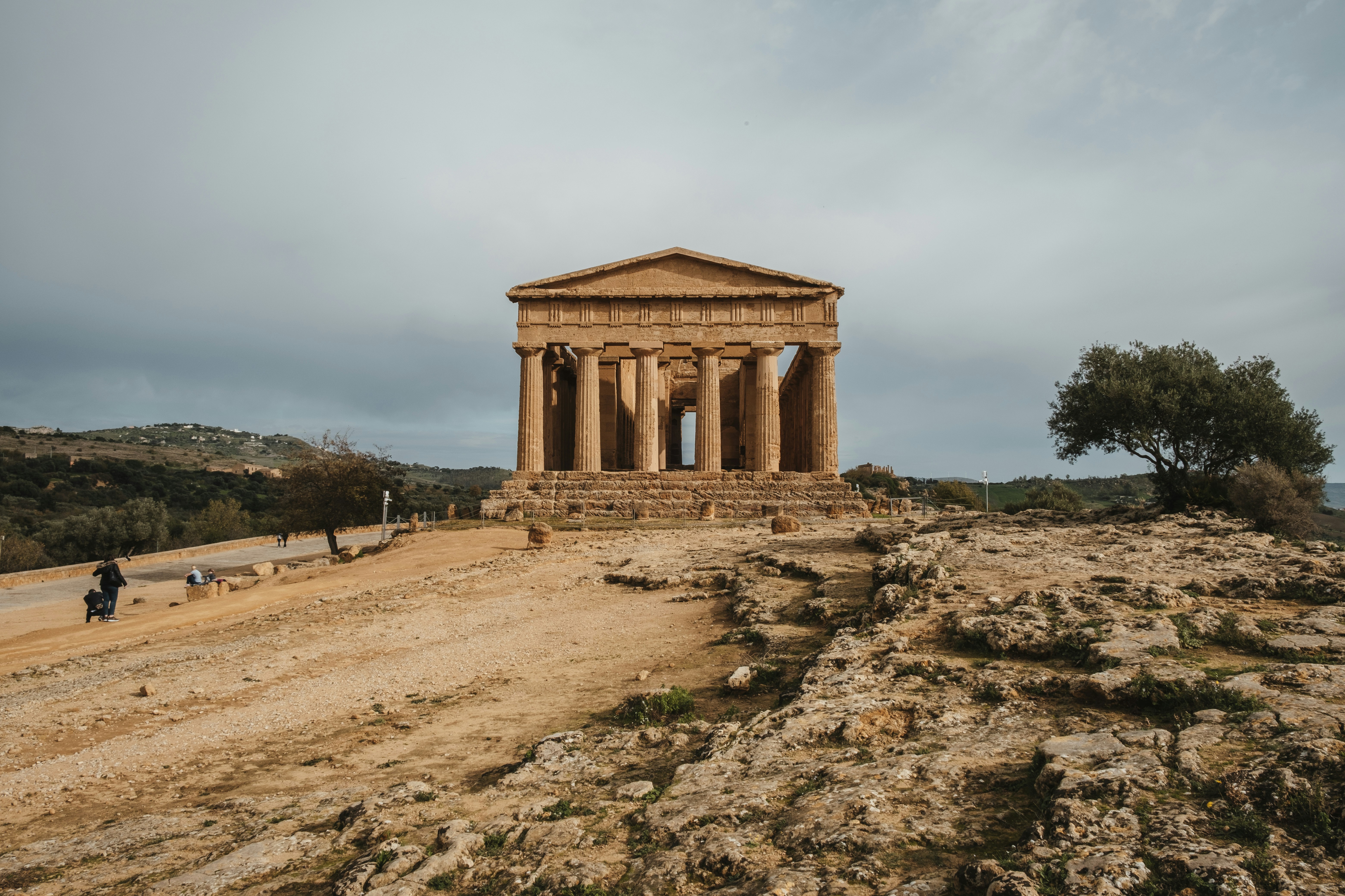 Ancient Greek temple with Doric columns stands amidst rocky terrain under a cloudy sky.