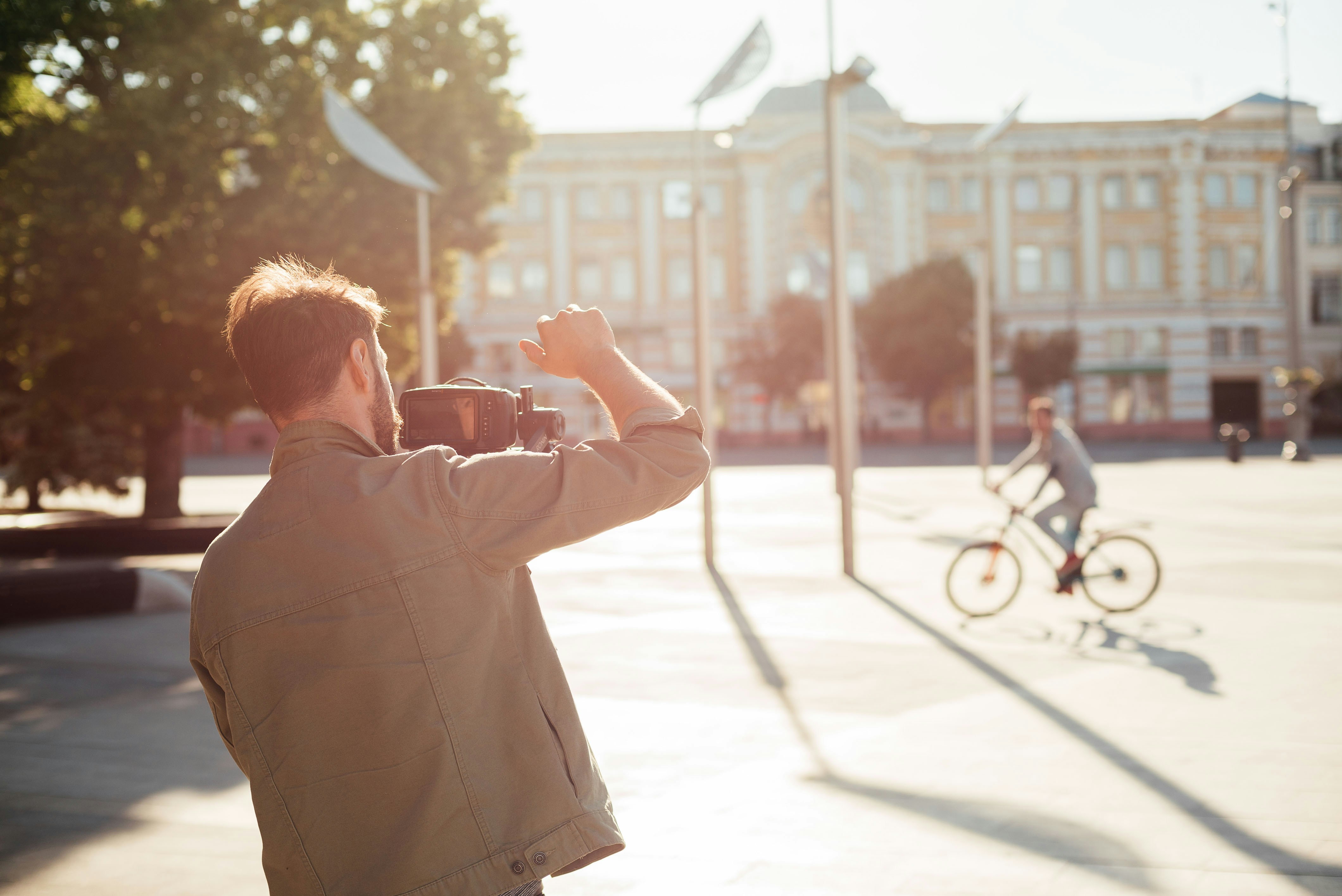 Man filming with a camera in a sunlit European square as a cyclist passes by.