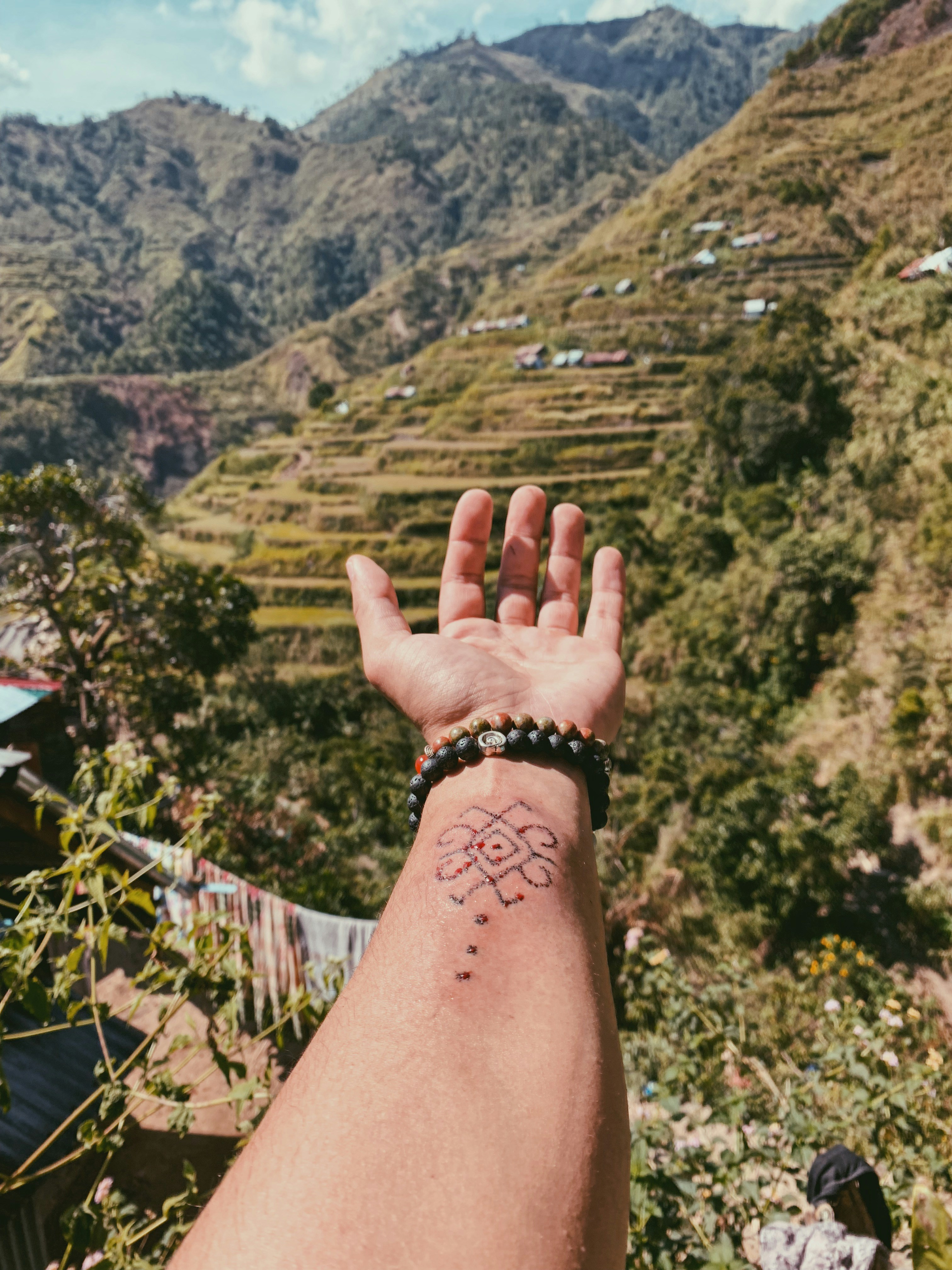 Hand in the foothills: tattoo, bracelet, and rising terraces under clear sky