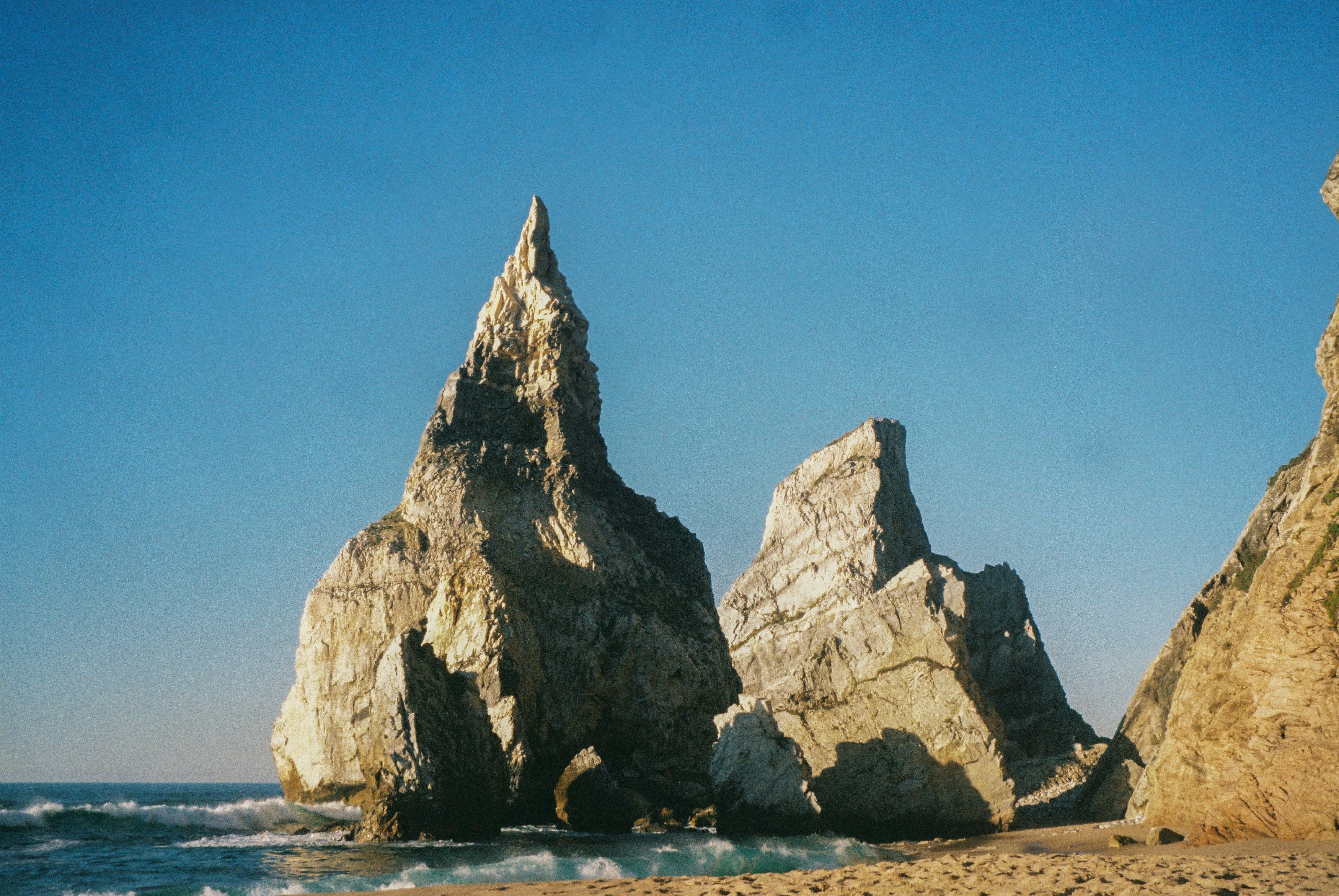 Three towering rock formations rise dramatically from the sandy beach, framed by a clear blue sky. The interplay of light and shadow accentuates their rugged textures.
