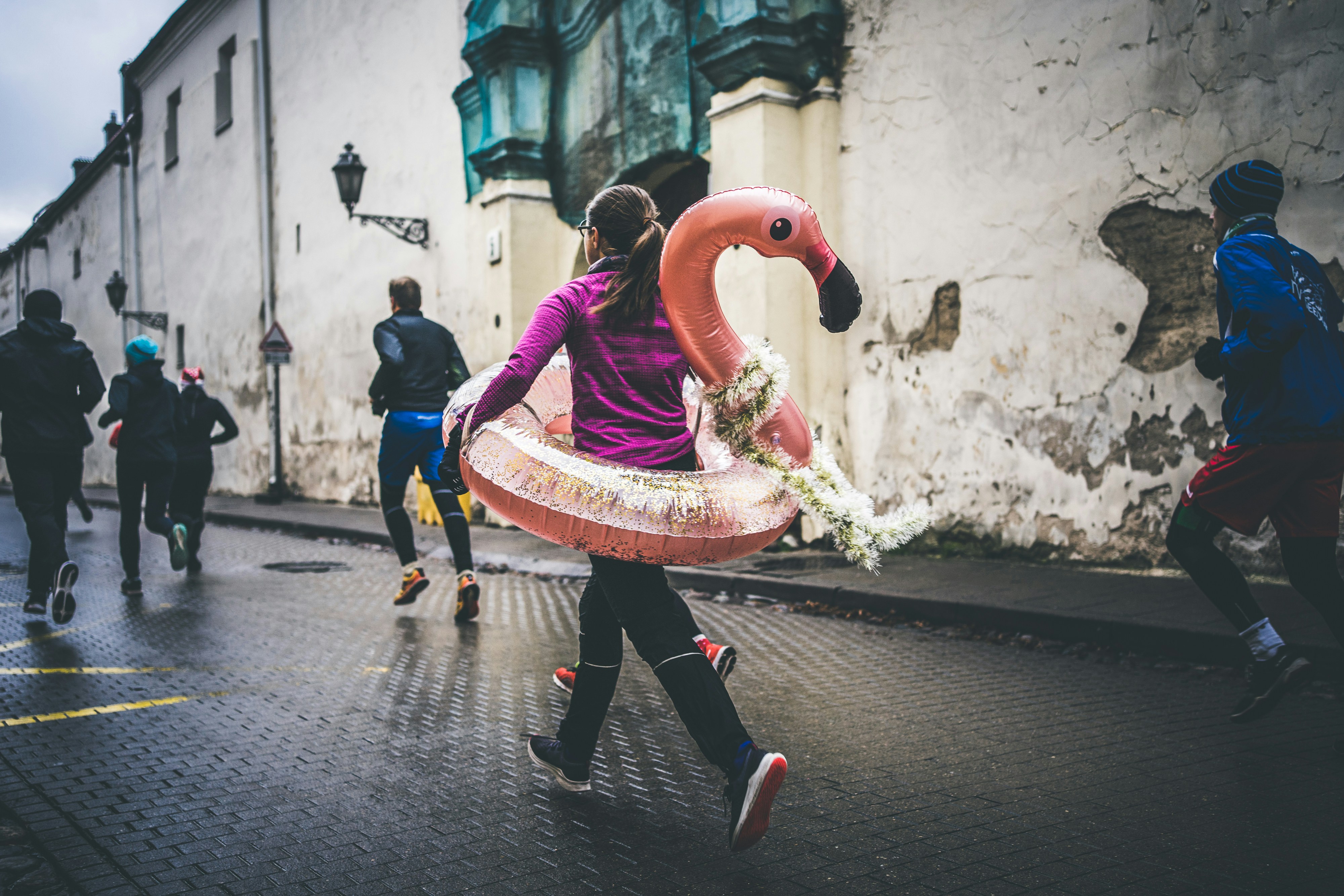 Person in a pink top carries a flamingo pool ring through a wet cobblestone street on a cloudy day.