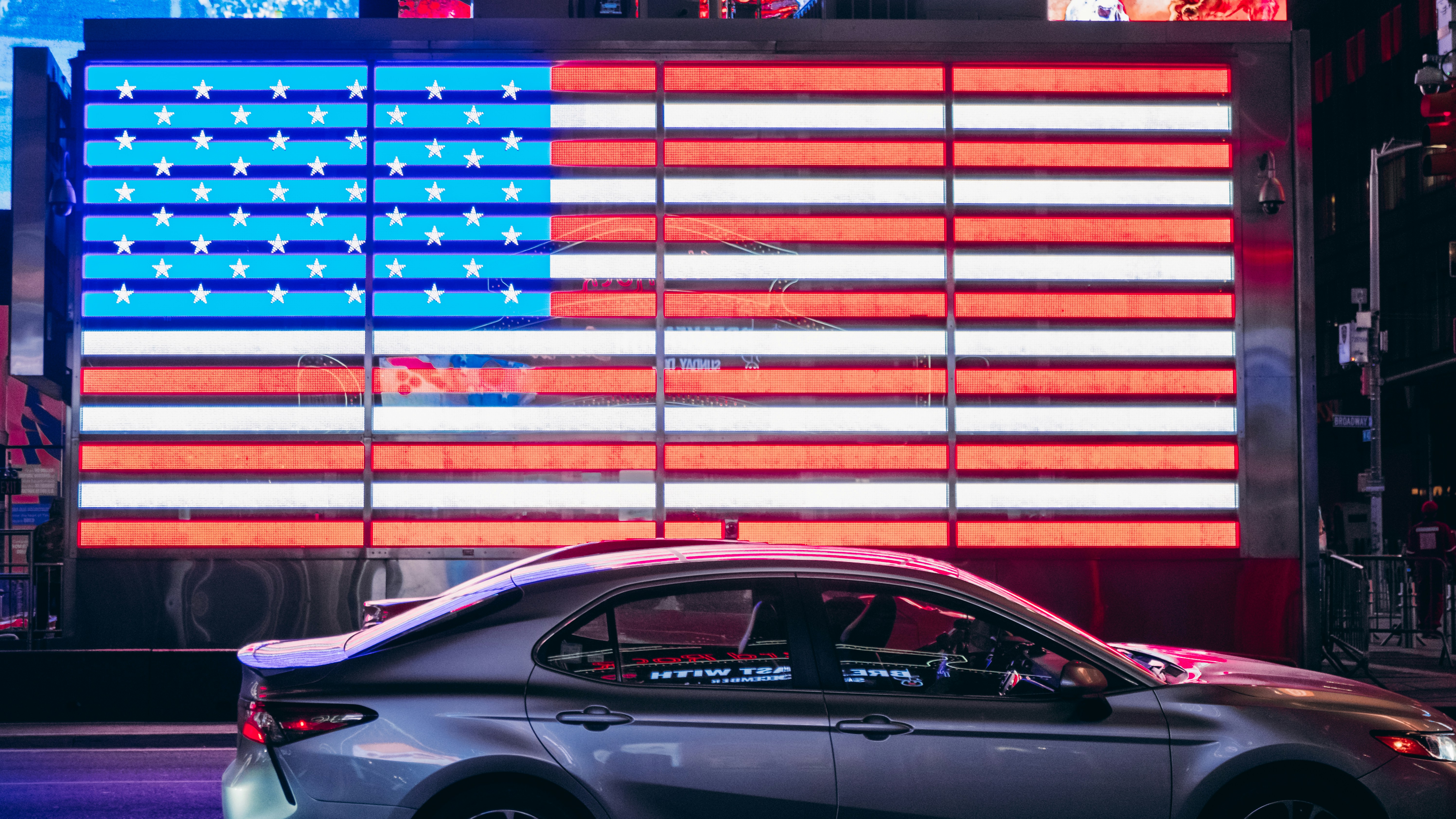 Chrome in Neon: A Silver Sedan Before a Giant LED American Flag