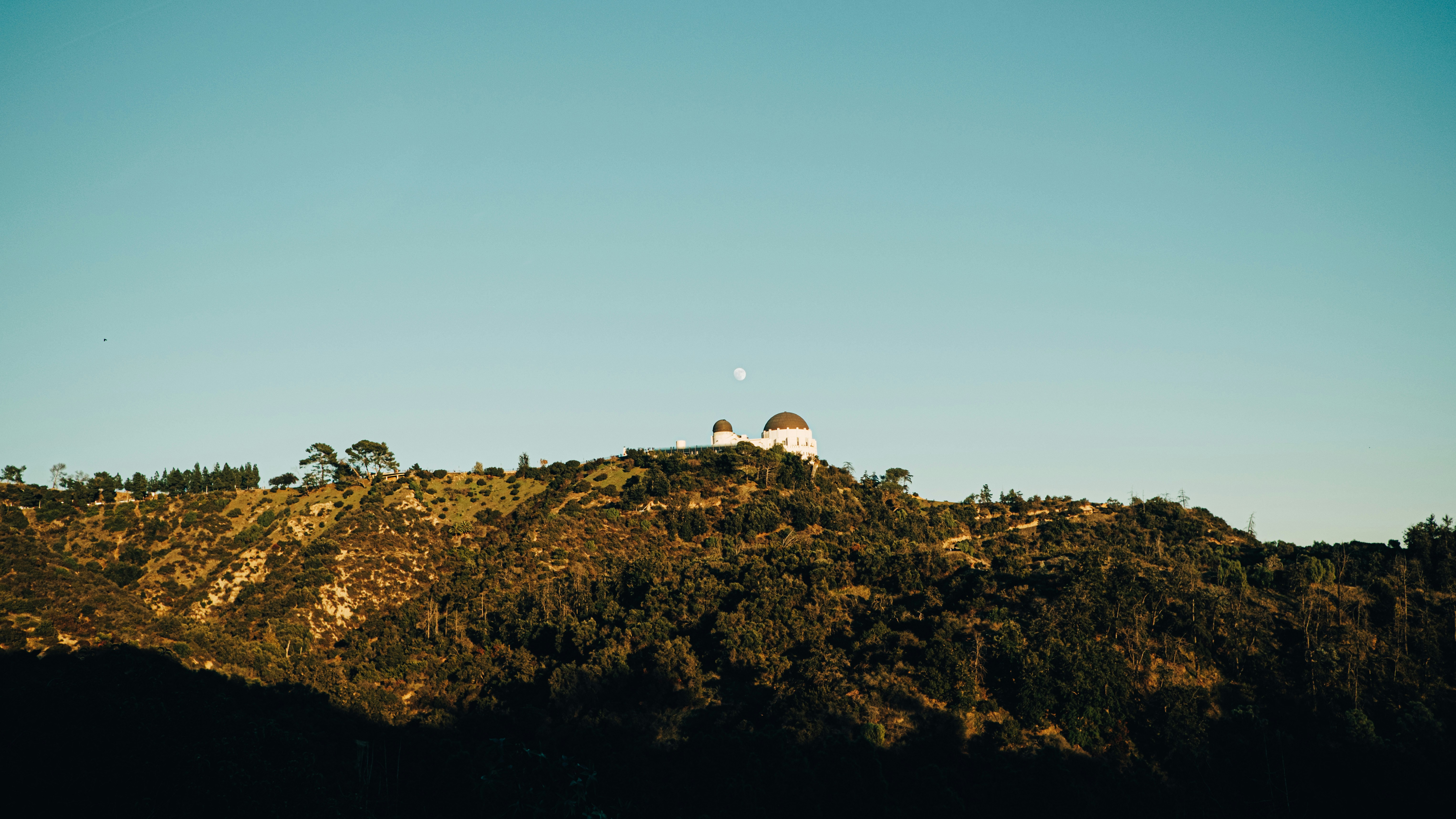 Griffith Observatory perched atop a hill, framed by lush greenery and a clear blue sky, with a crescent moon rising in the background.