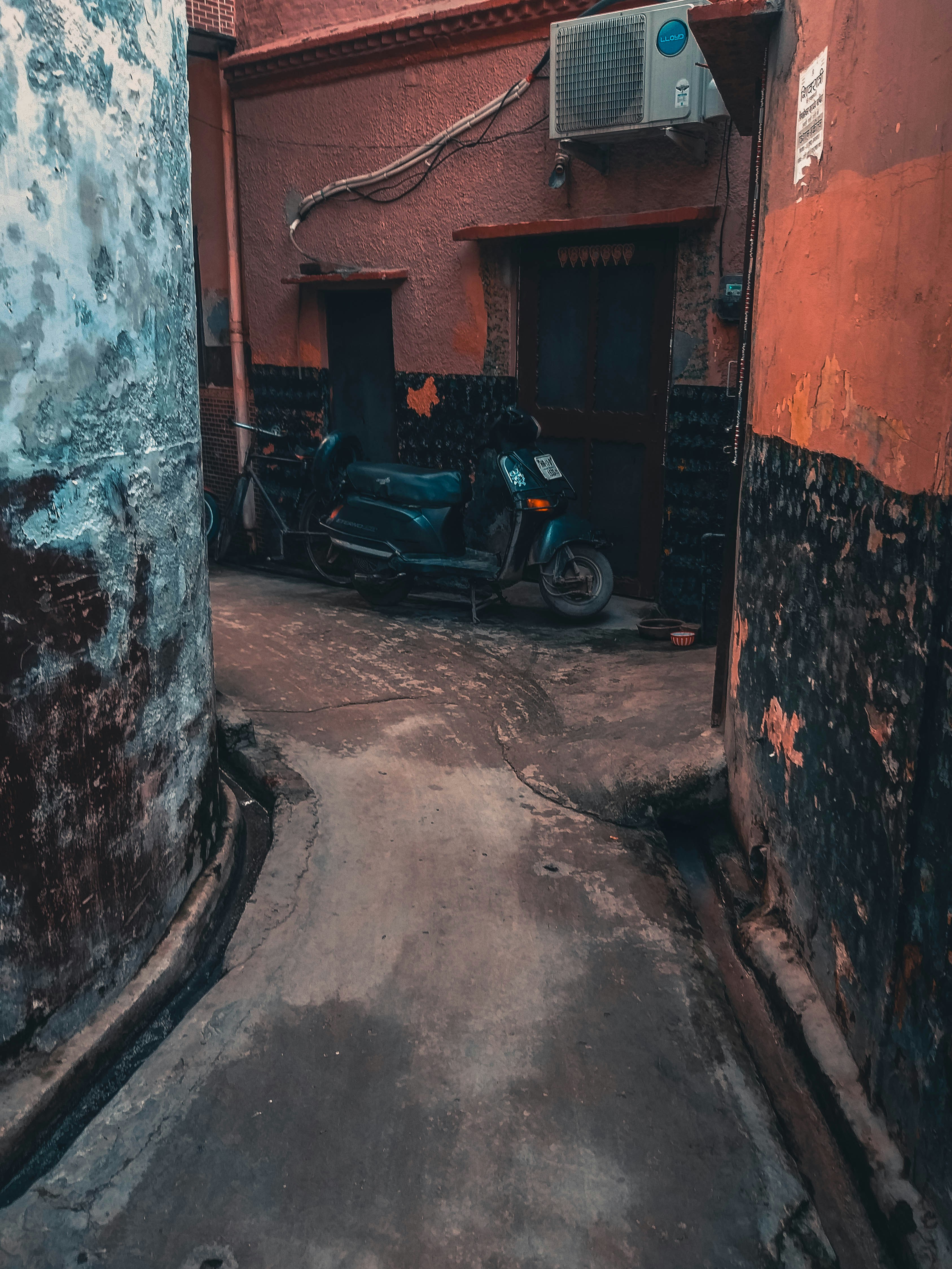 Narrow alleyway in an urban setting, featuring a parked motorcycle and textured walls with peeling paint. The scene captures the essence of city life.