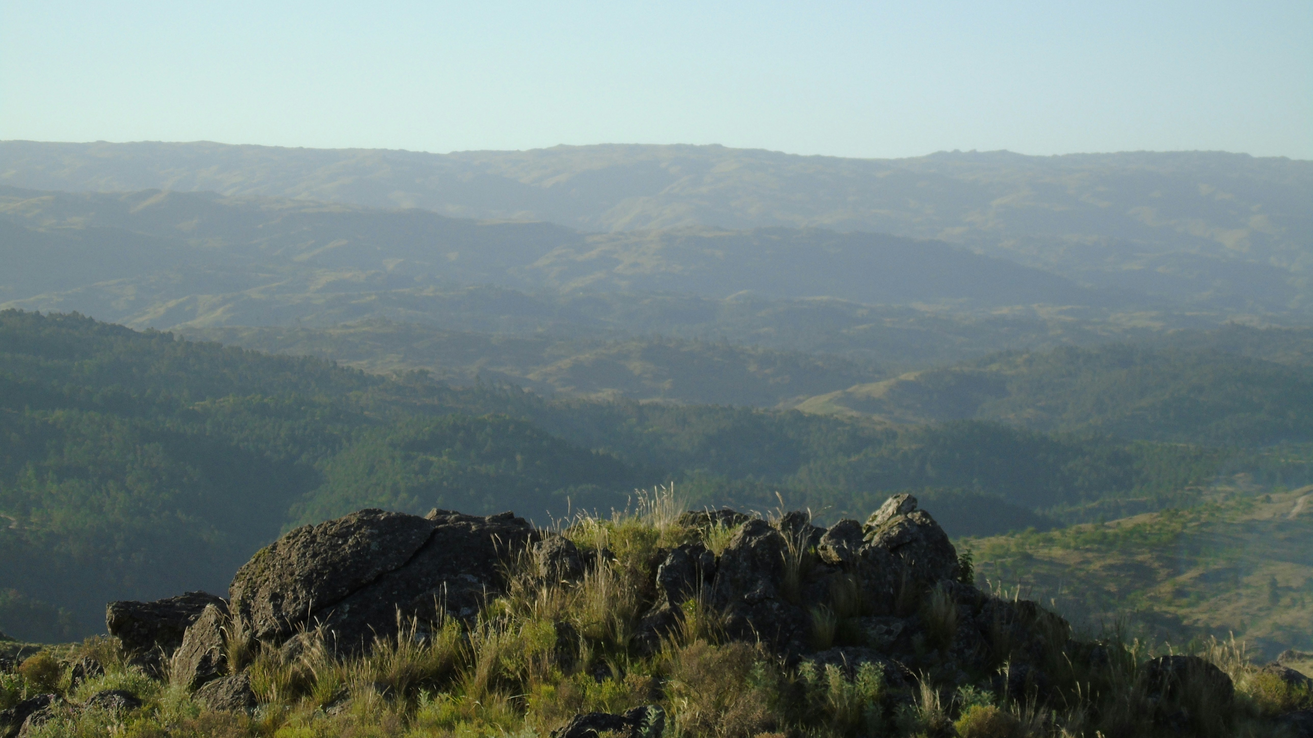 Lush green hills stretch across the horizon, framed by rocky outcrops under a clear sky. The scene evokes a sense of tranquility and connection with nature.