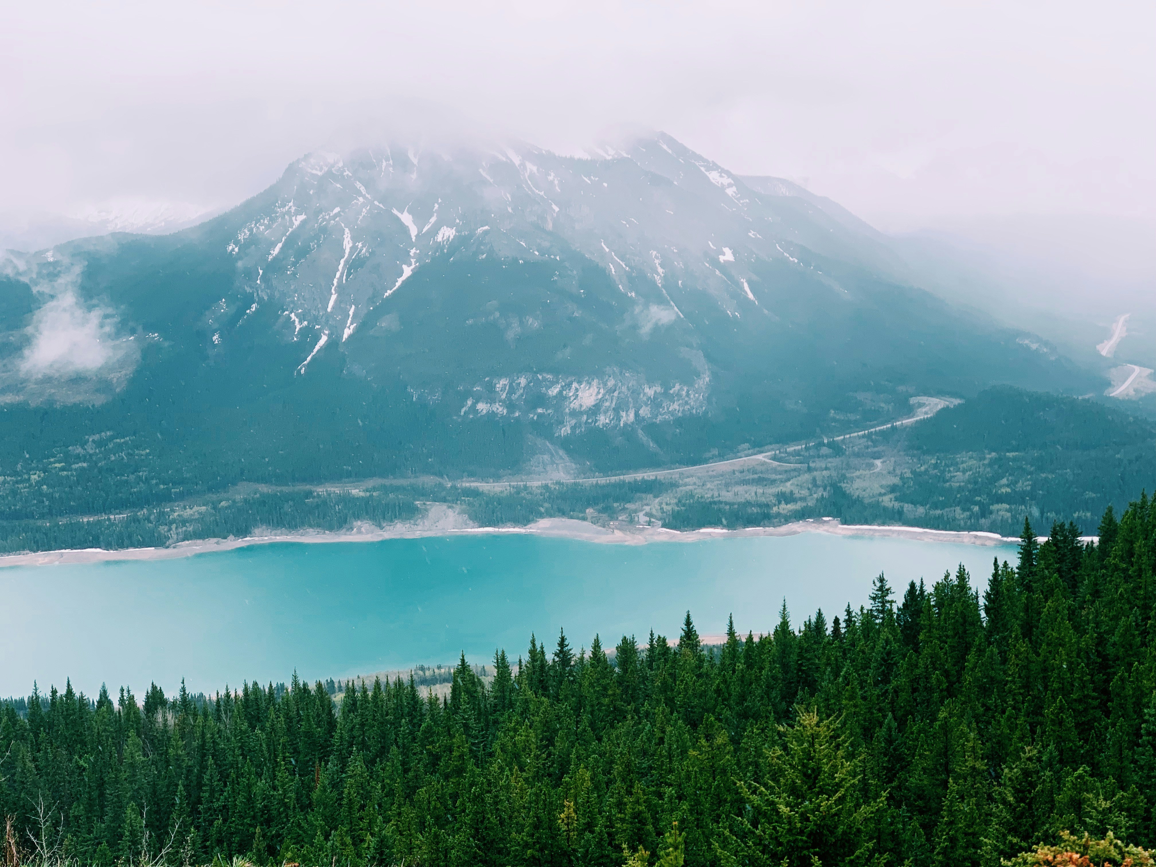 Snow-dusted mountain shrouded in mist above a turquoise lake with lush evergreen forest.