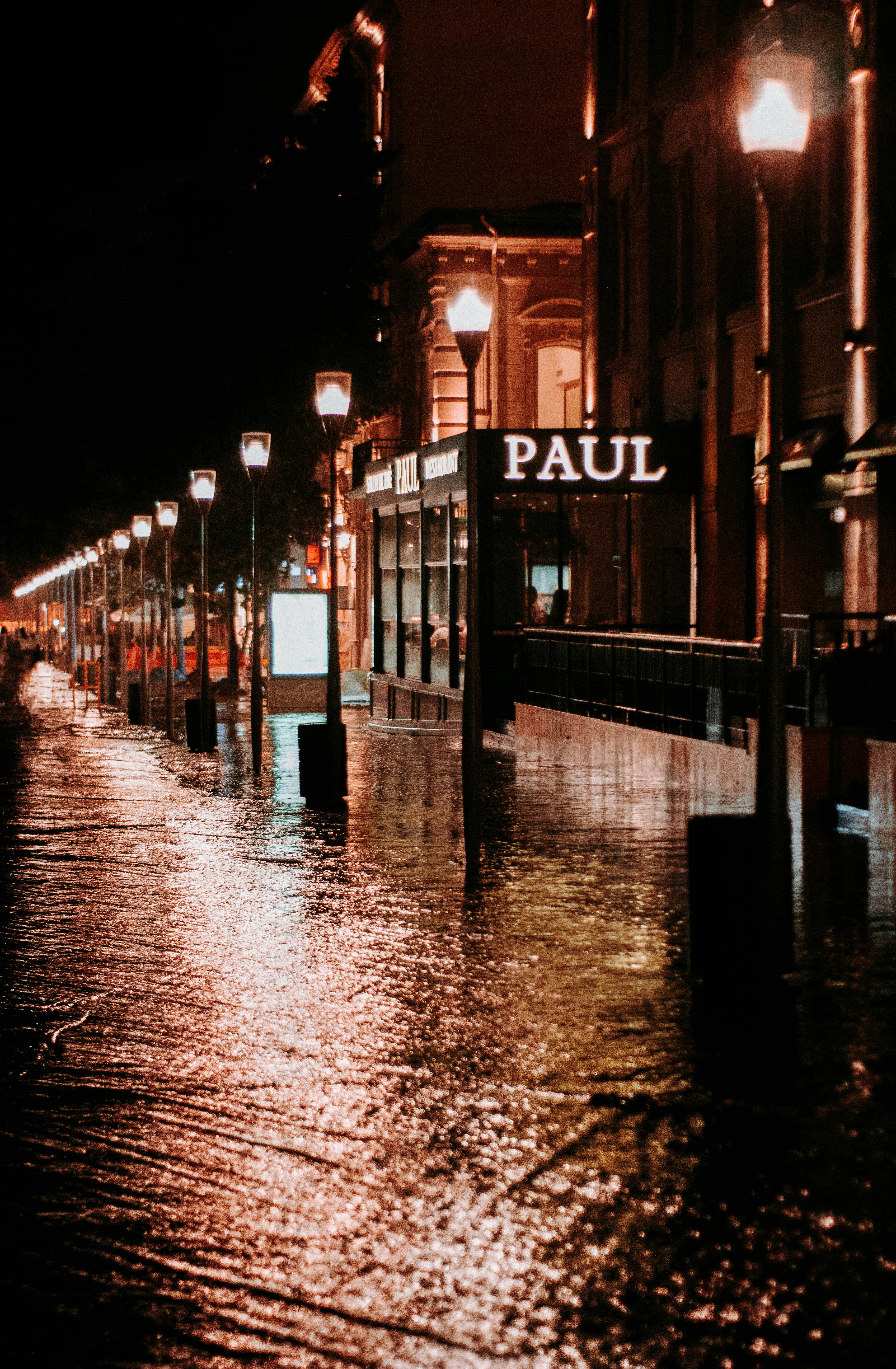 Street illuminated by lampposts reflecting on a waterlogged pavement near a restaurant named PAUL.