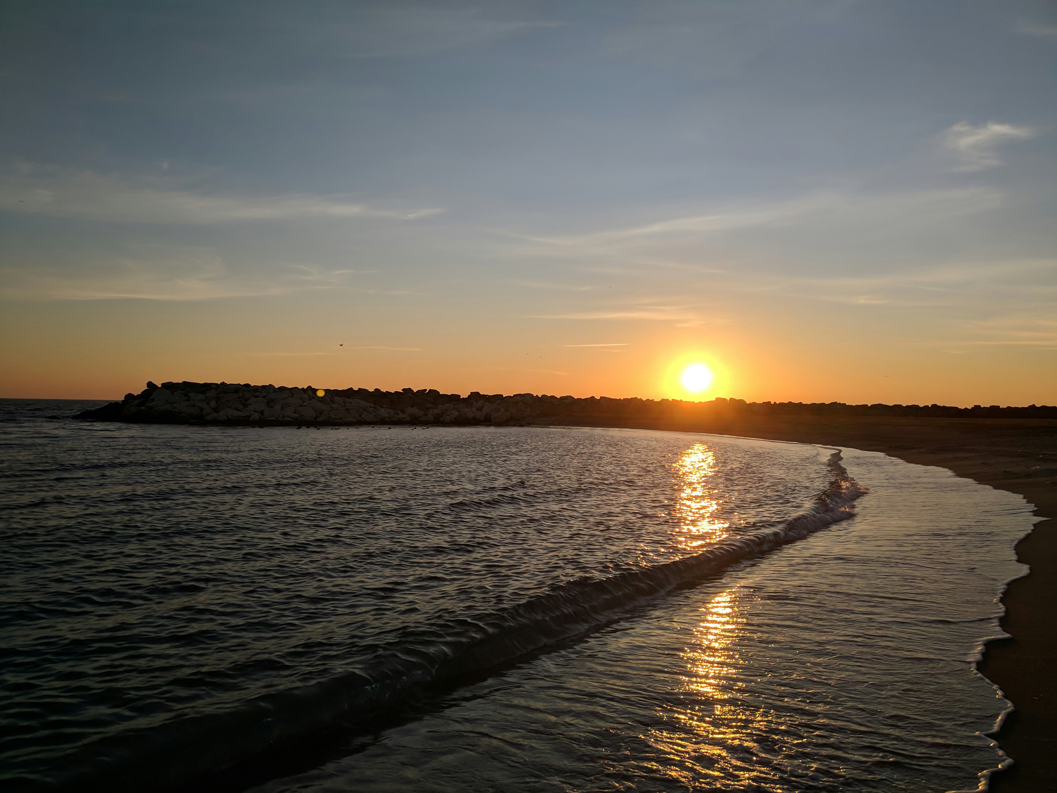 Golden sun setting over a tranquil beach, casting reflections on gentle waves. The shoreline curves gracefully, inviting contemplation.