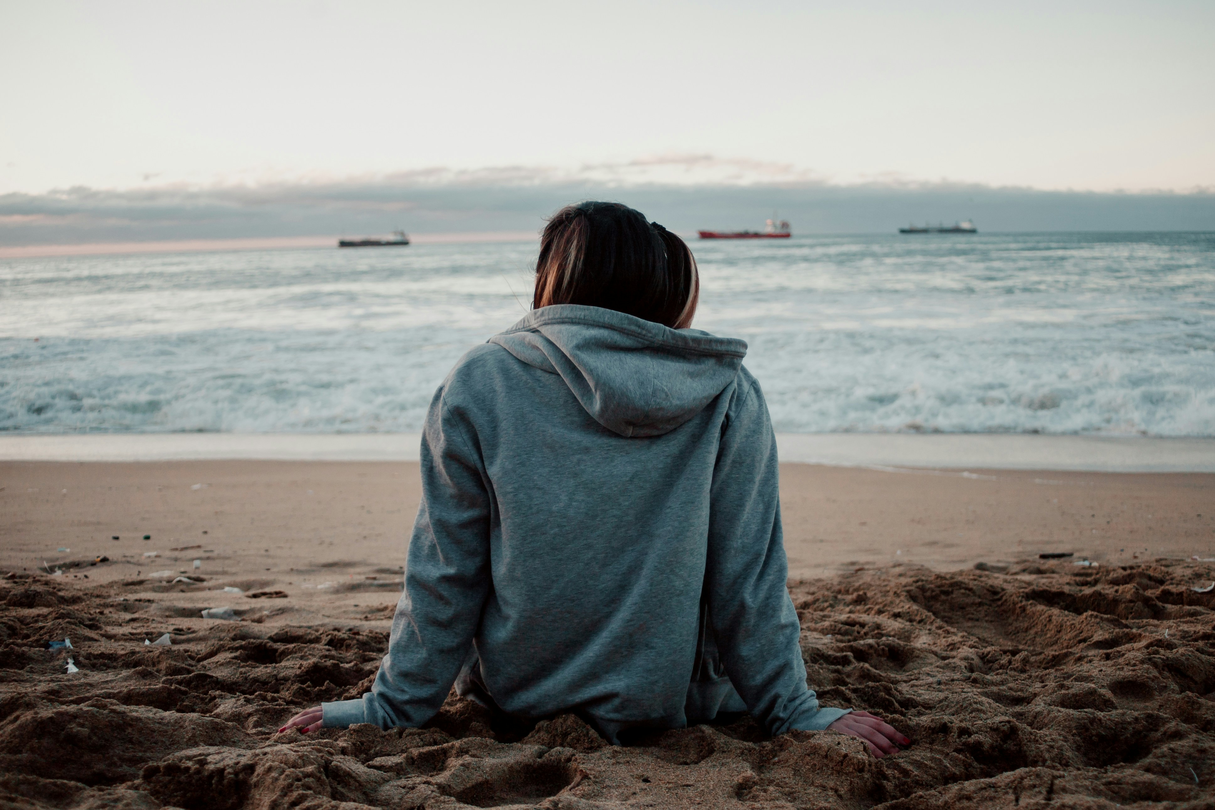Person in a gray hoodie sits on a sandy beach facing the ocean with distant ships on the horizon.