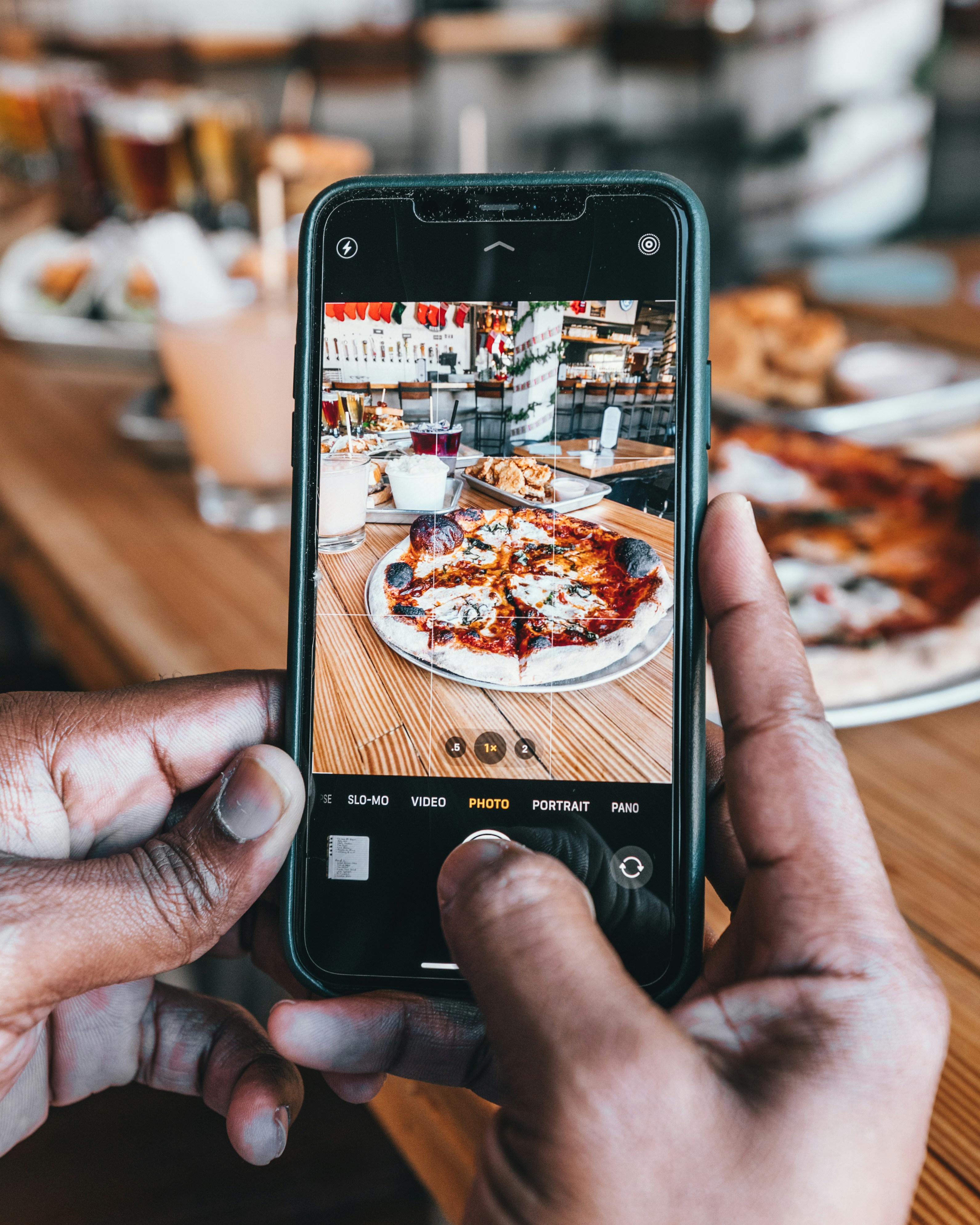 A hand holds a smartphone, capturing a delicious pizza on a wooden table amidst a vibrant dining scene. Various dishes and drinks are visible in the background.