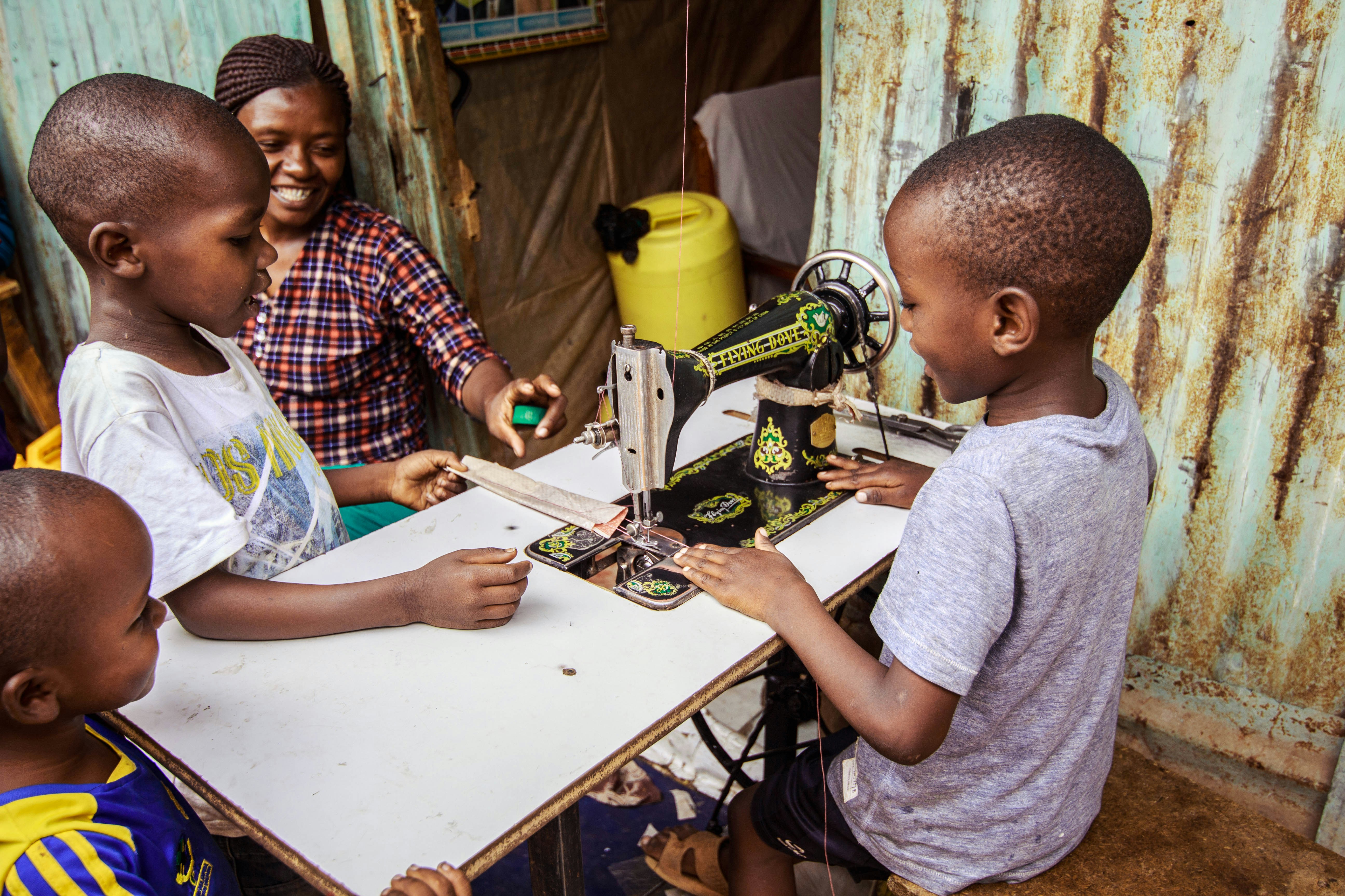 Children learning to use a vintage sewing machine under the guidance of an adult in a rustic setting.