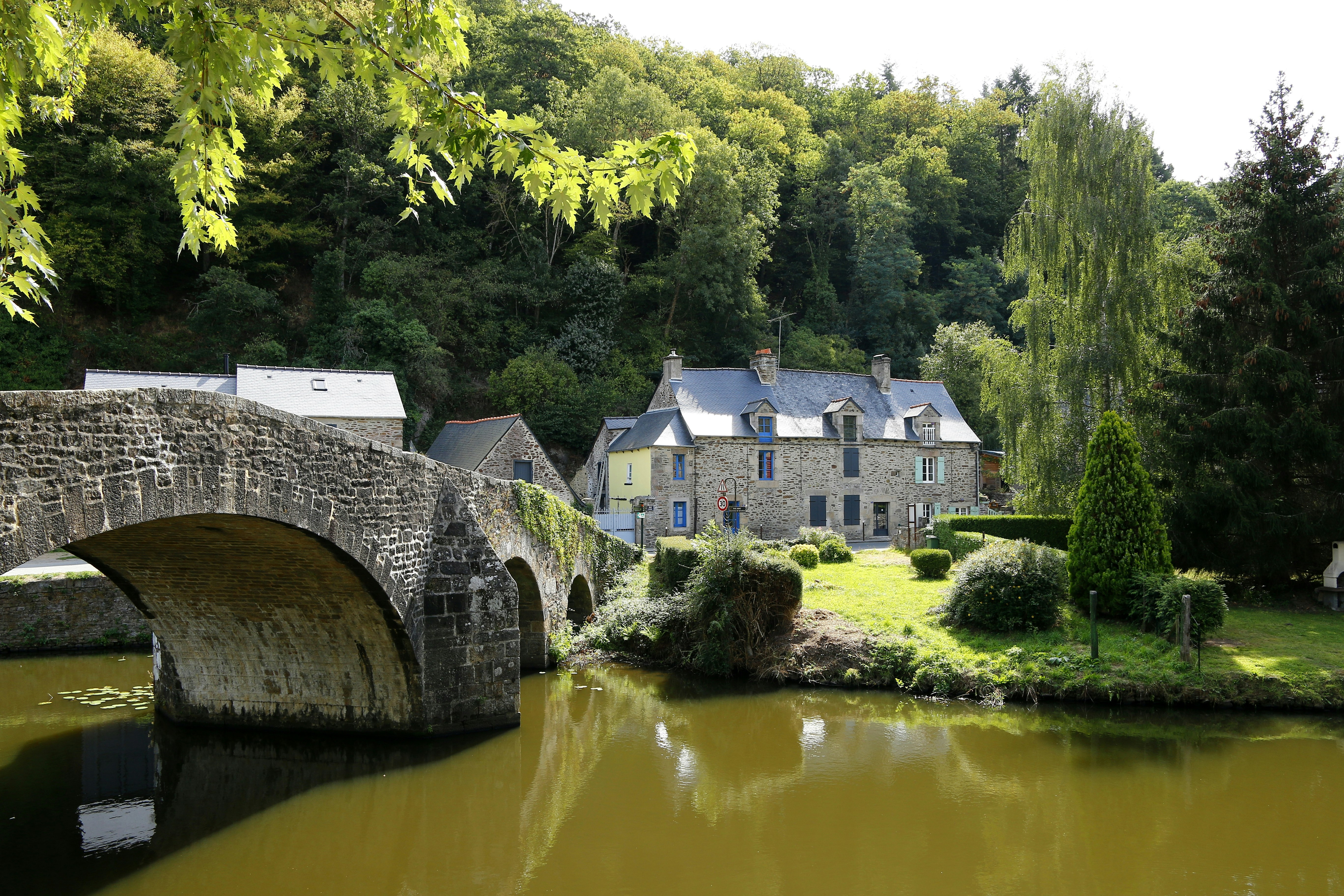 Historic stone bridge arches over a calm river beside a quaint house surrounded by lush greenery.