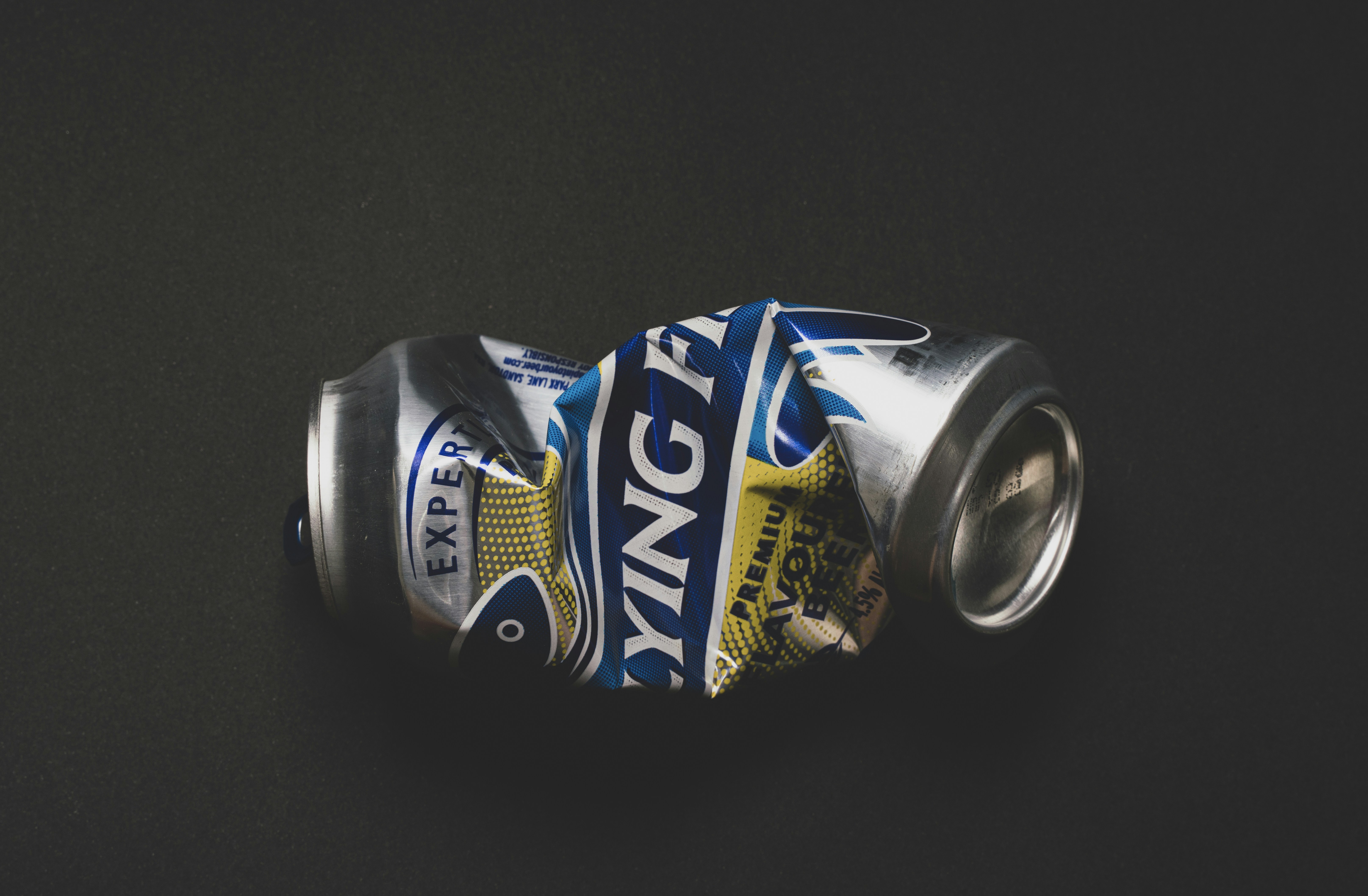 Crumpled soda can resting on a dark surface, showcasing vibrant colors and intricate designs. The image highlights themes of waste and consumerism.