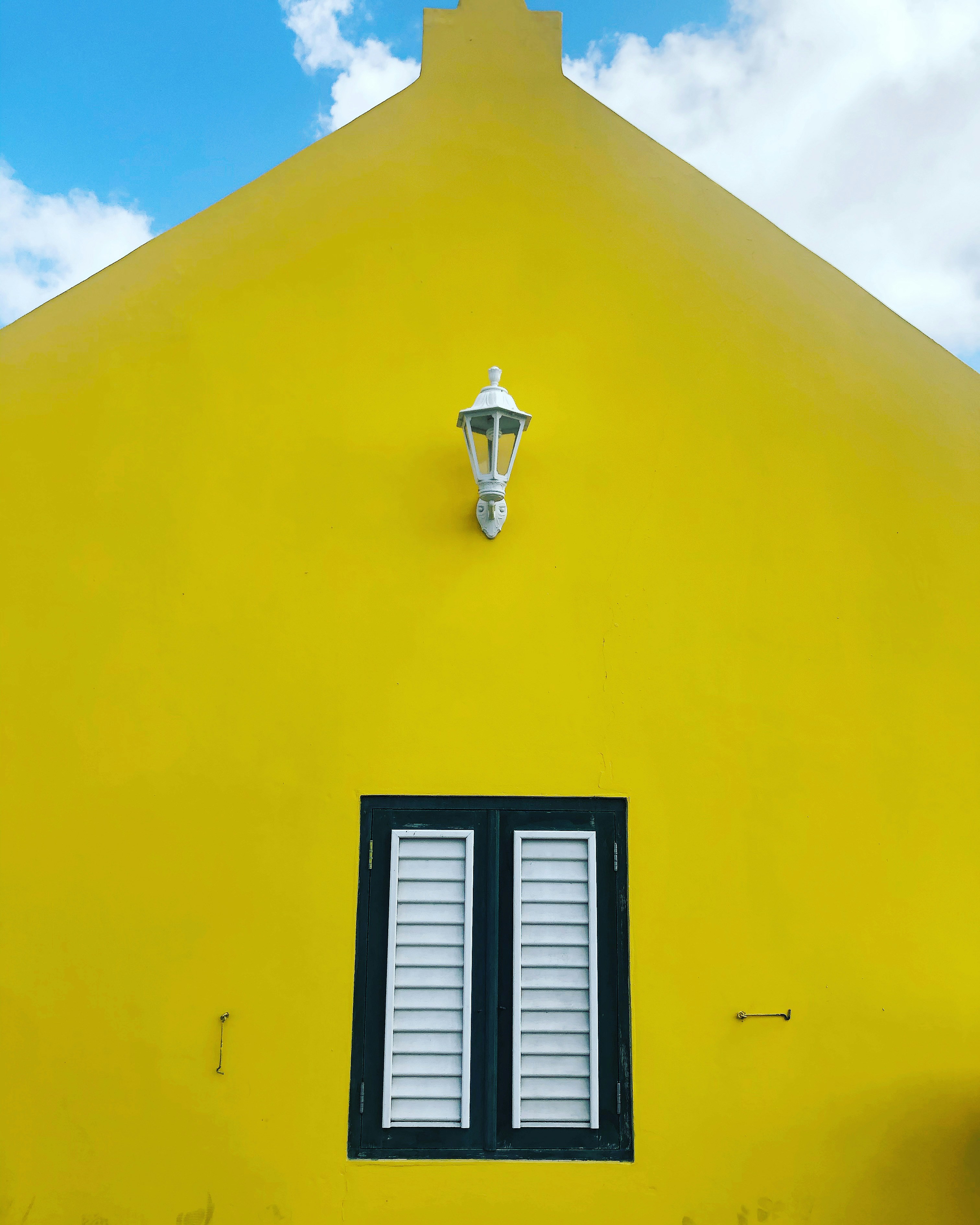 Bright yellow wall featuring a black-framed window with shutters and a vintage lamp above. The clean lines and bold color create a striking visual impact.