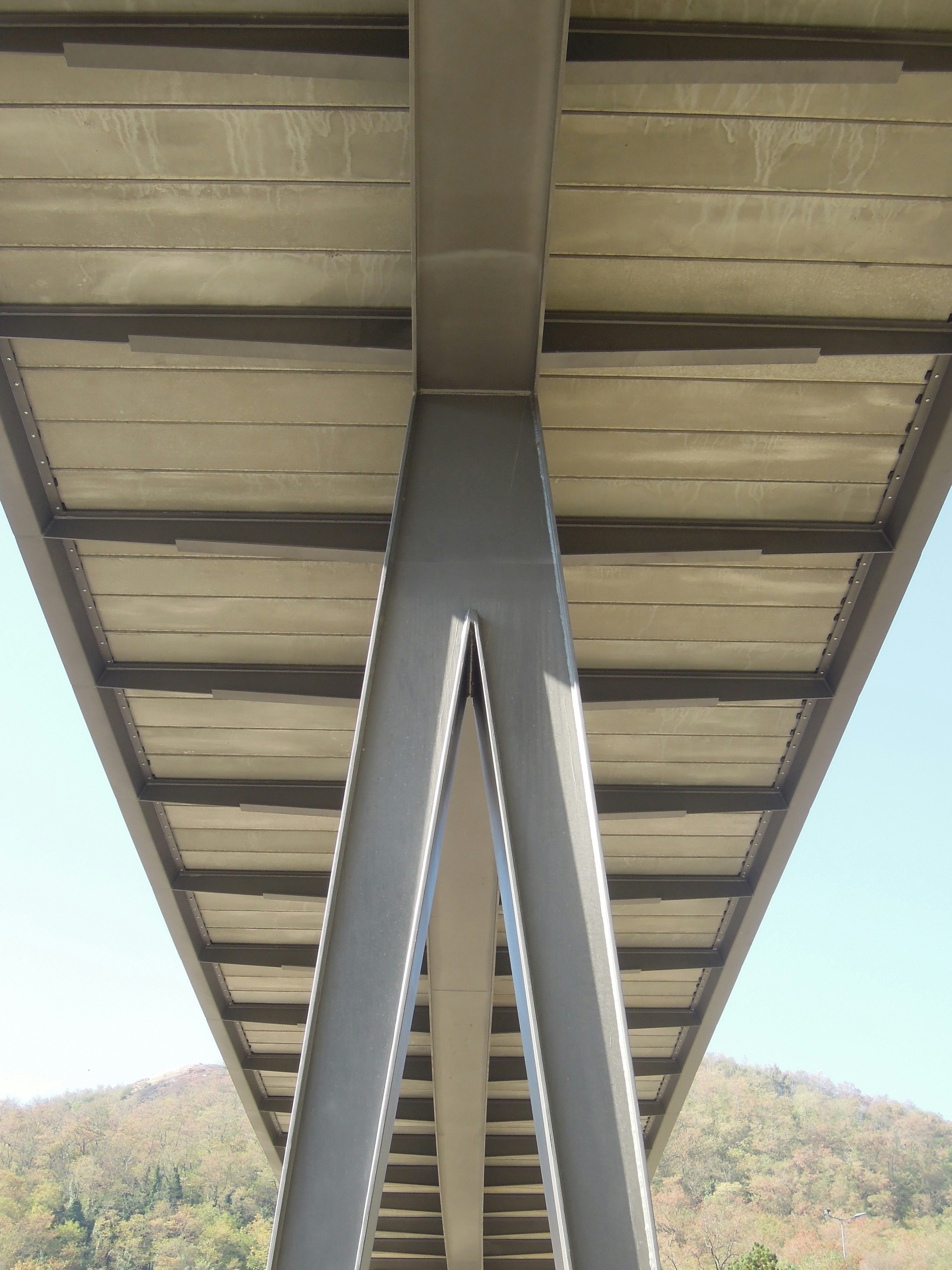 View from beneath a modern bridge showcasing its intricate support beams and textured underside against a backdrop of rolling hills.