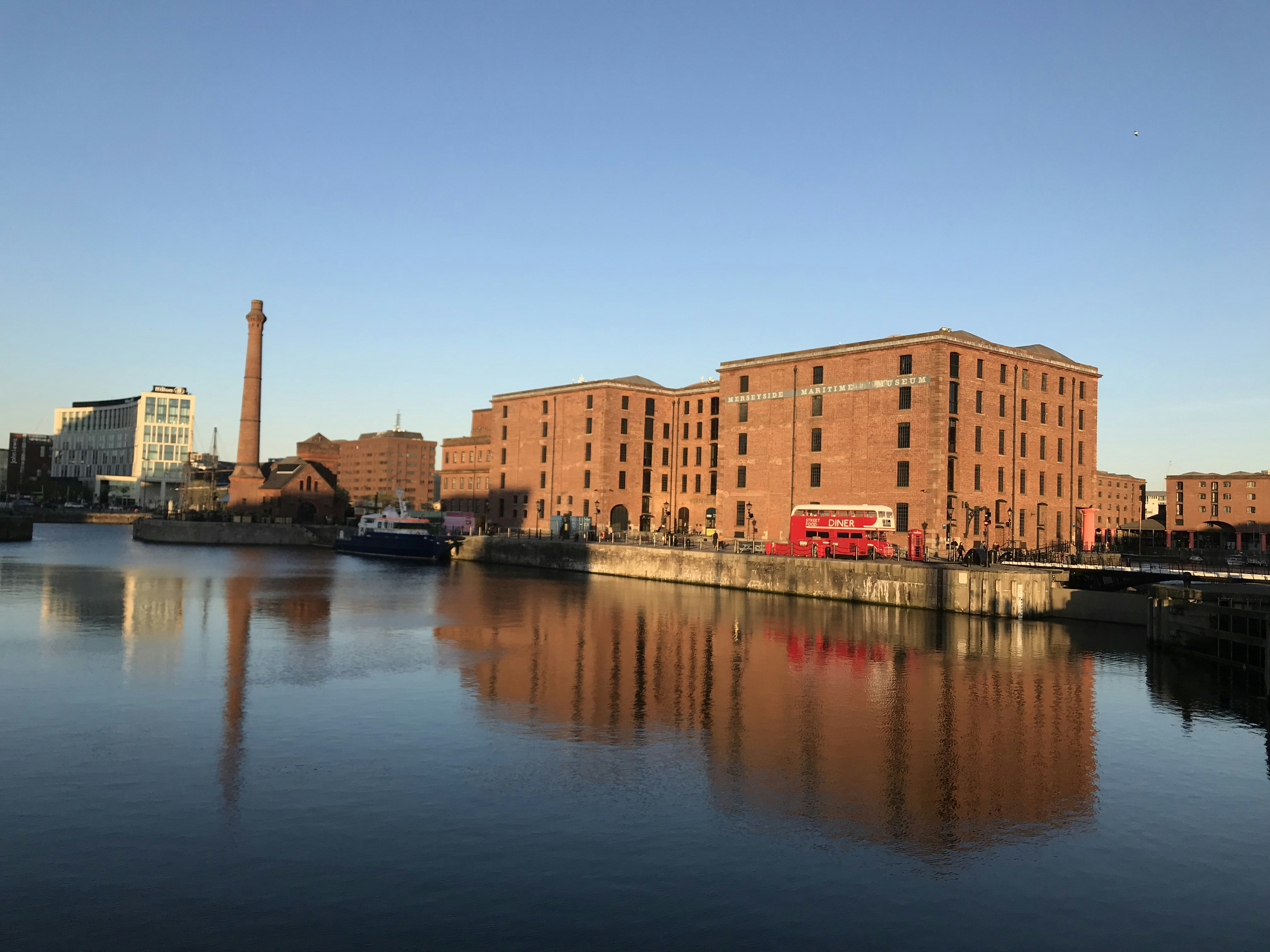 Historic brick warehouses reflected in calm waters under a clear blue sky.