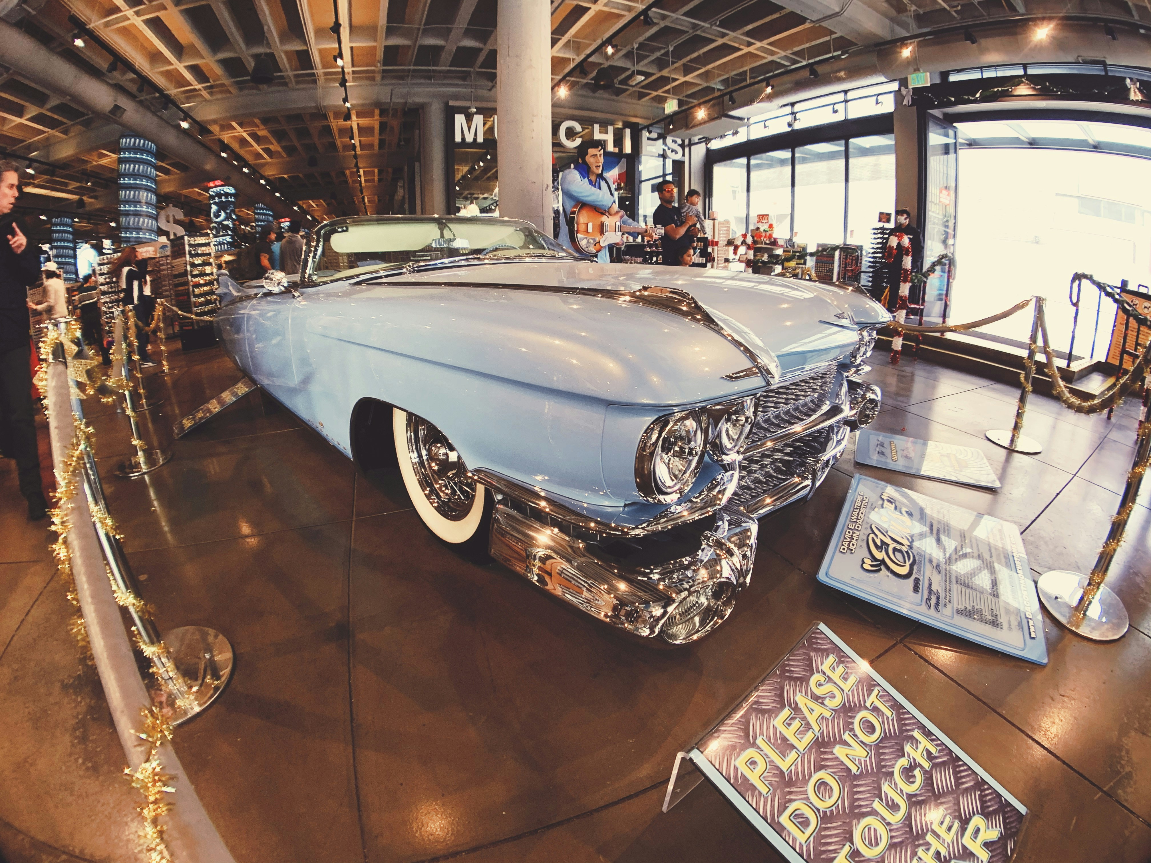 Vintage light blue car with chrome detailing showcased indoors, surrounded by onlookers and festive decorations.