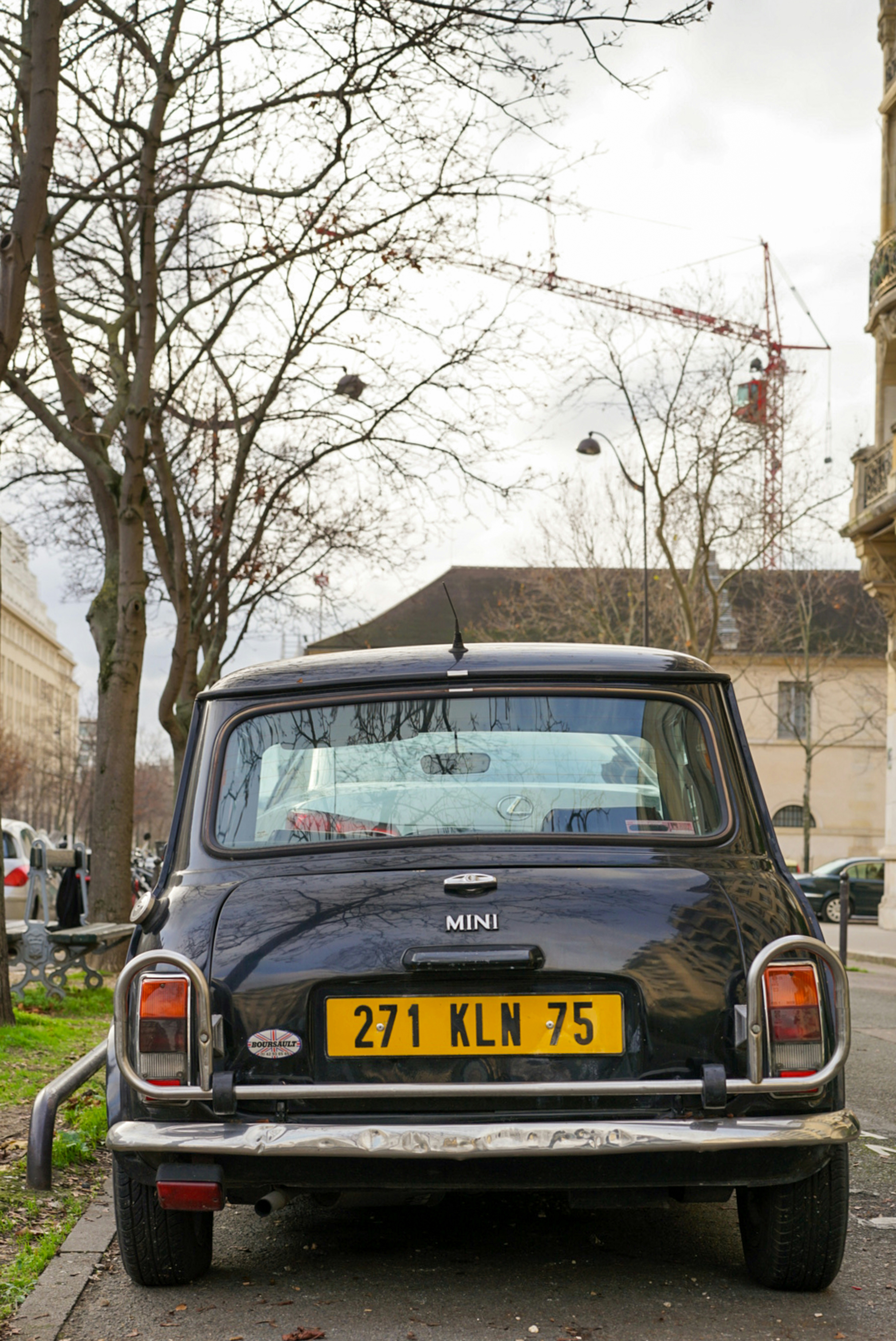 Classic black Mini parked on a city street, surrounded by bare trees and urban architecture. A construction crane looms in the background.
