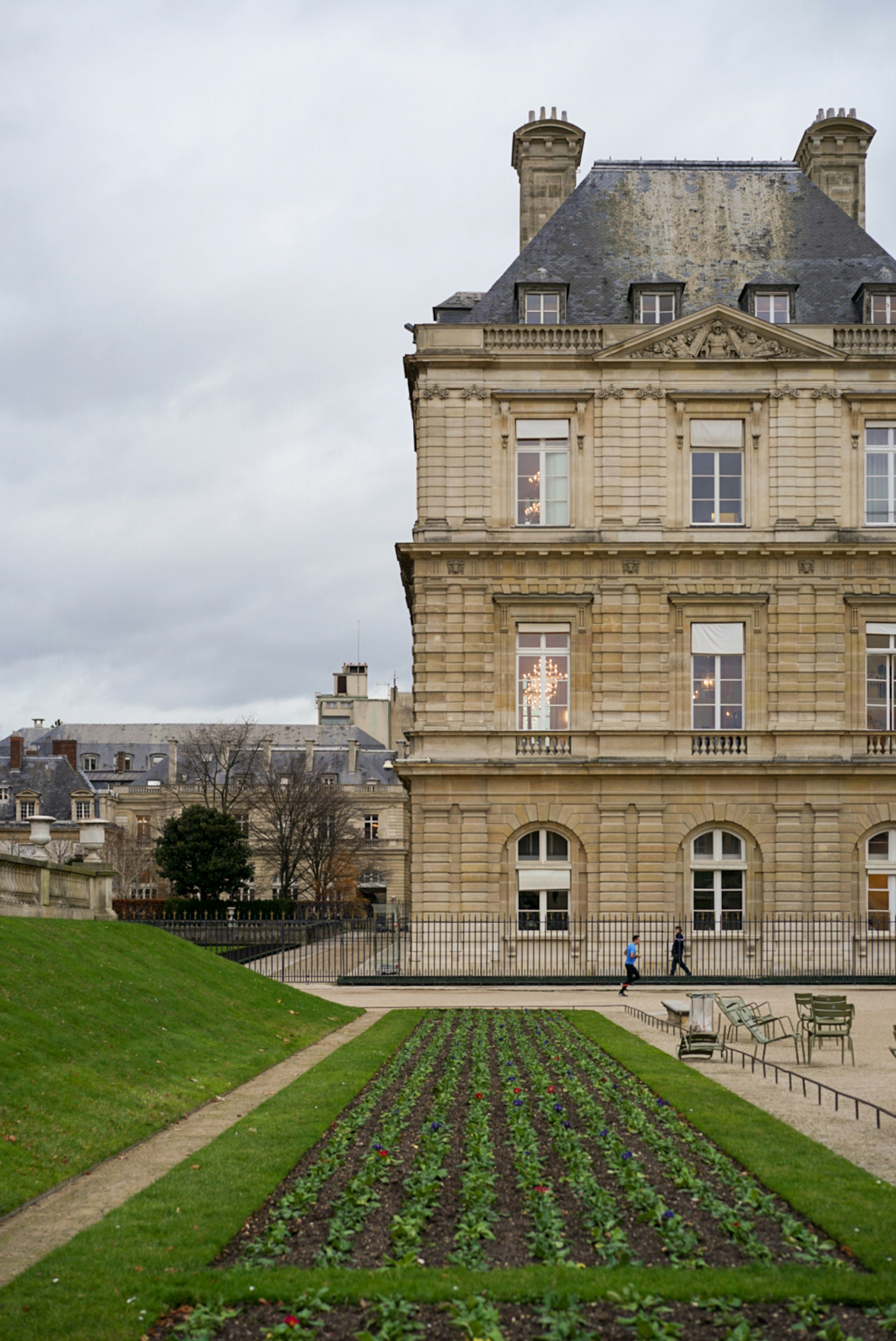 Elegant historic building framed by manicured gardens, with two figures strolling along the path. The scene captures a blend of architecture and nature.