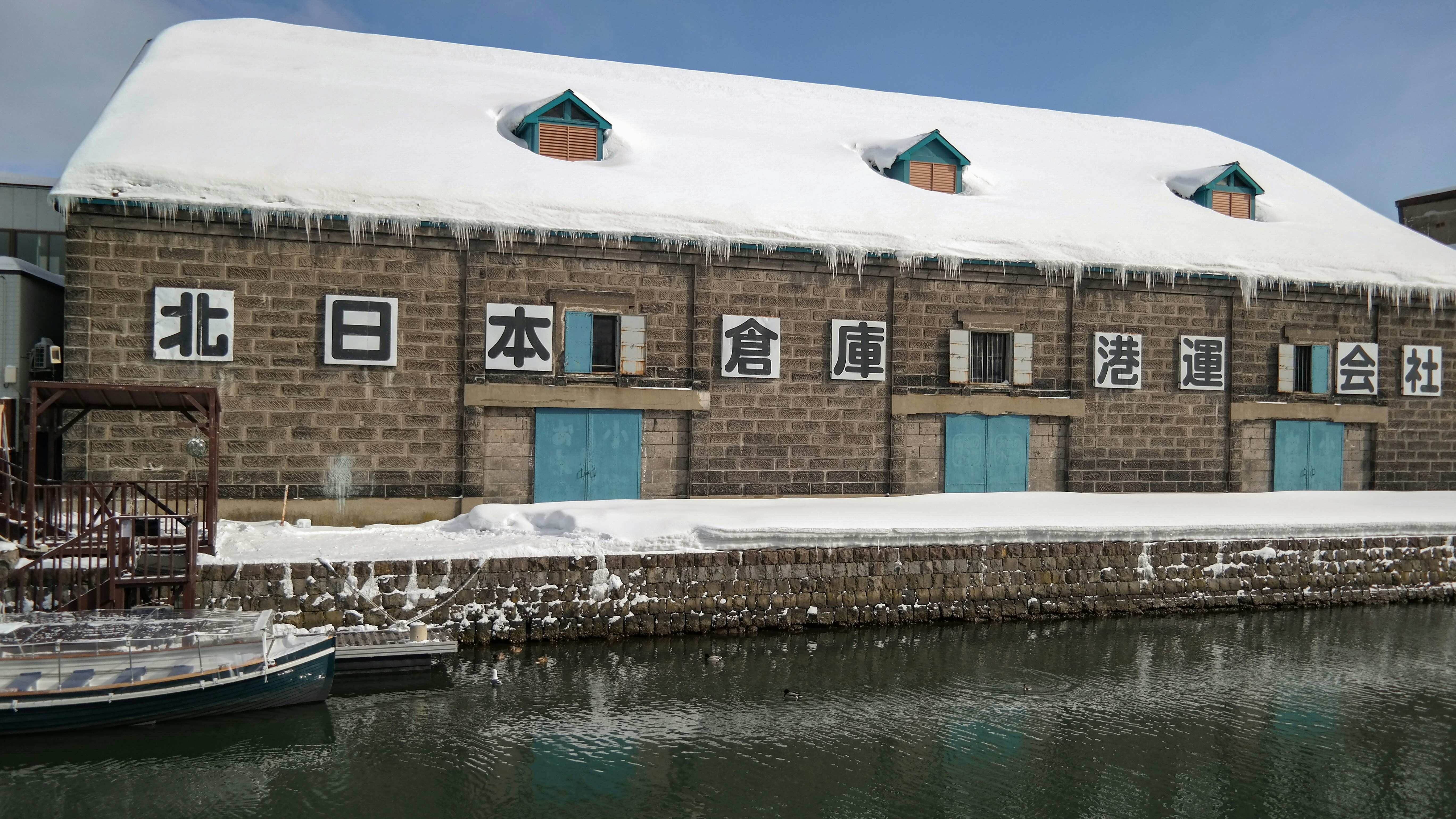 Snow-clad brick warehouses line a calm canal, their faded blue doors contrasting with the snow and water. Bold kanji signage anchors the façade and lends a strong sense of place.