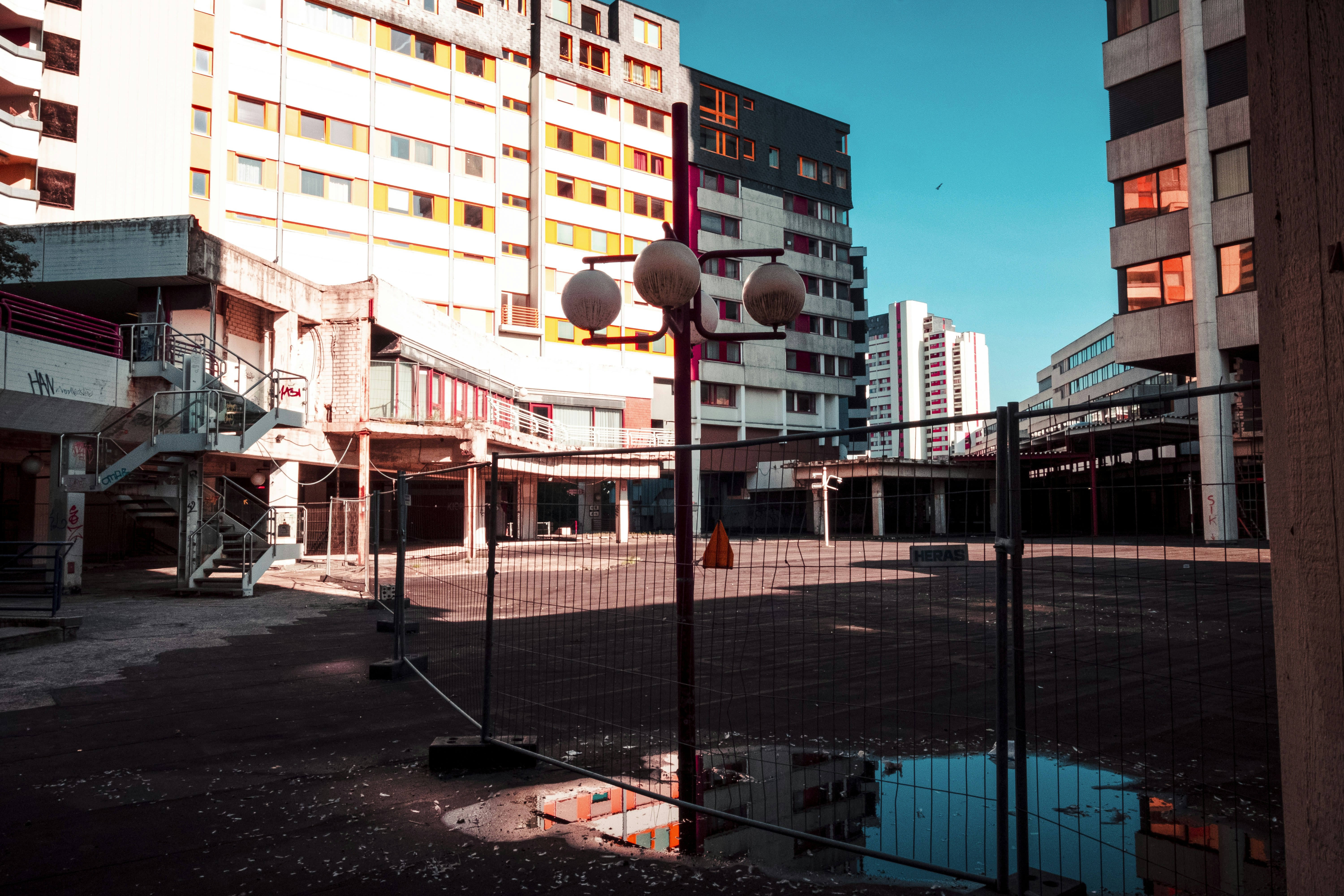Deserted urban courtyard surrounded by modernist buildings, featuring a reflective puddle and stark fencing. The scene conveys a sense of desolation and architectural contrast.