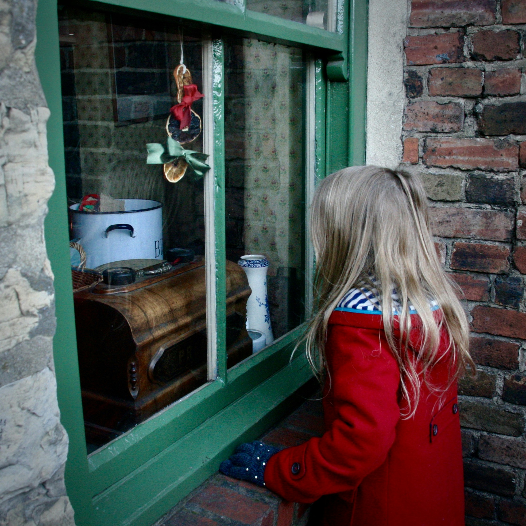 Child in a red coat gazing through a green-framed window, revealing a glimpse of a vintage interior adorned with nostalgic decor.