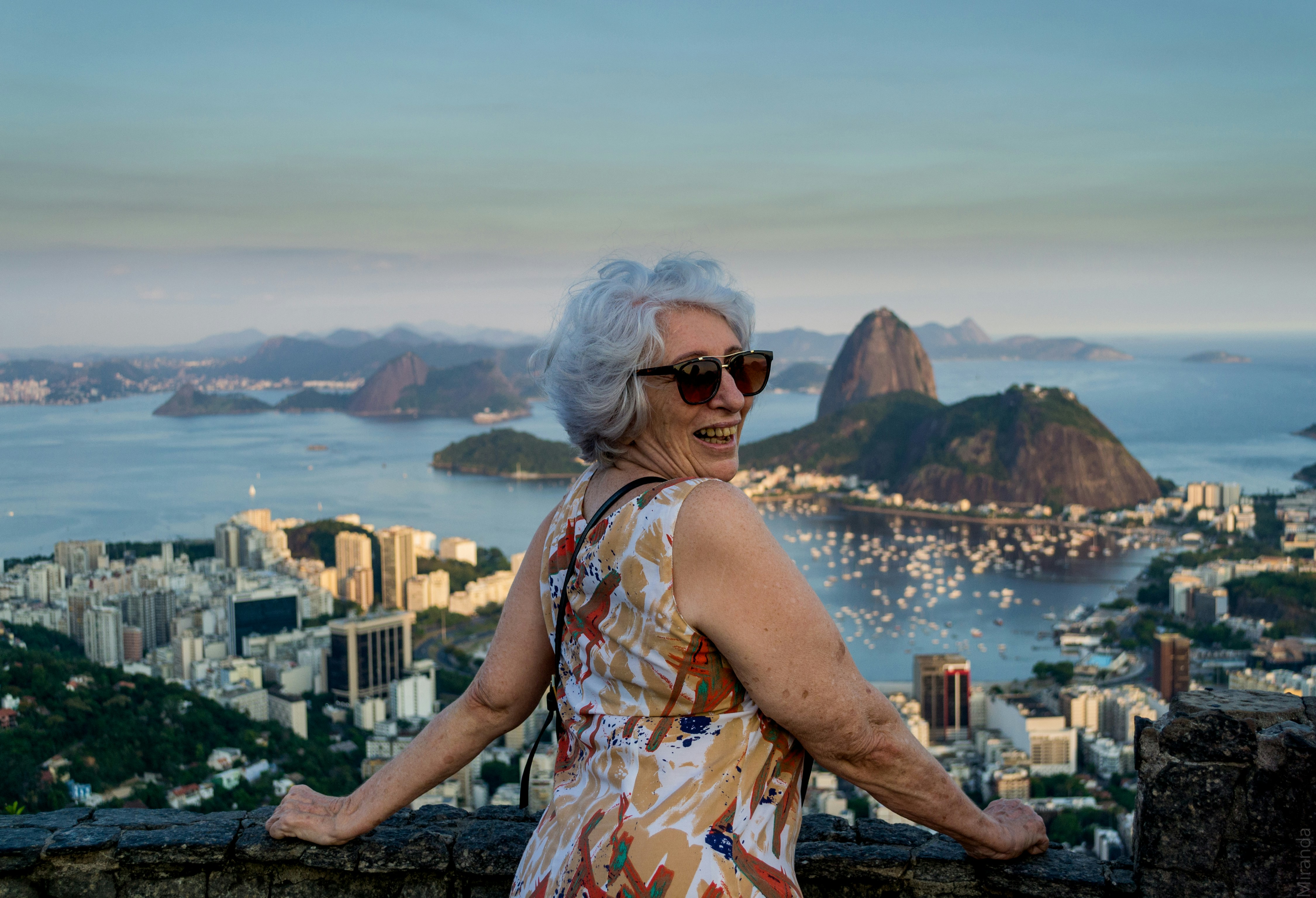 Elderly woman in a colorful dress smiles while leaning on a stone rail with Rio de Janeiro's skyline and Sugarloaf Mountain in the background.