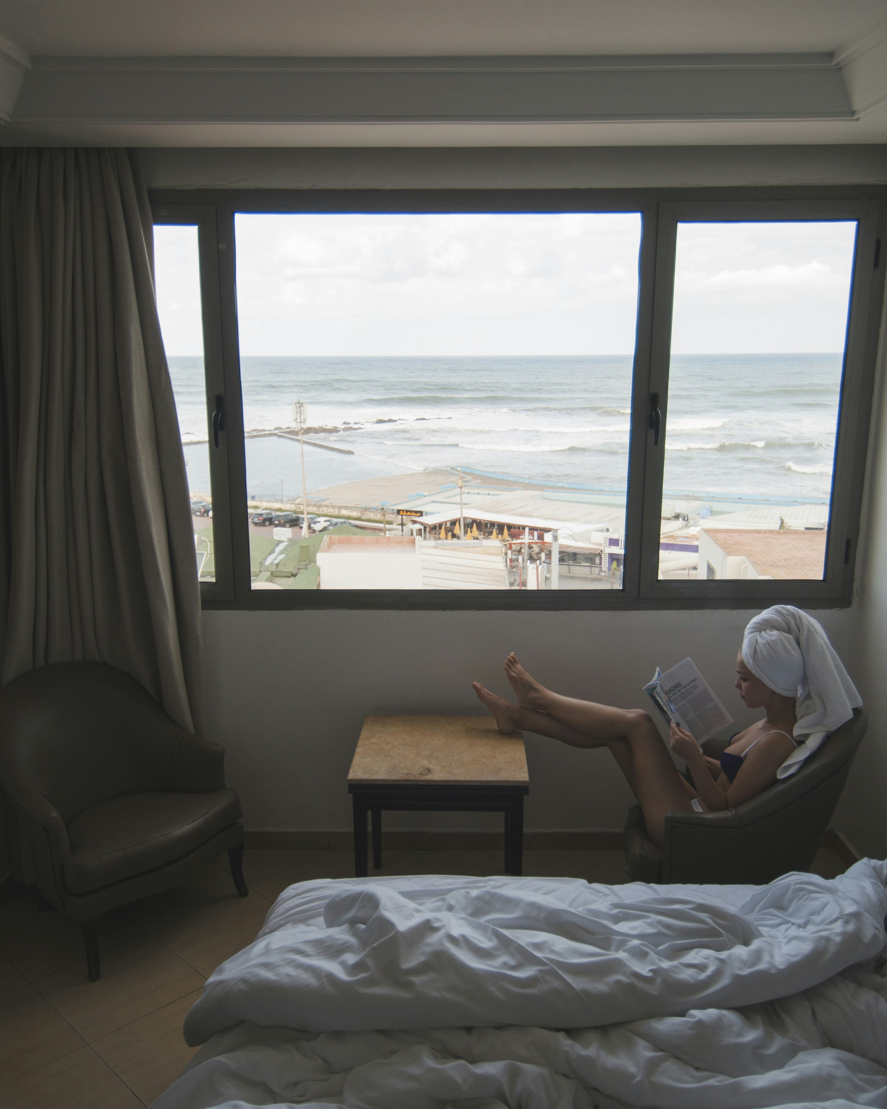 Woman relaxing in a cozy room, engrossed in a book while gazing out at the ocean view through large windows.