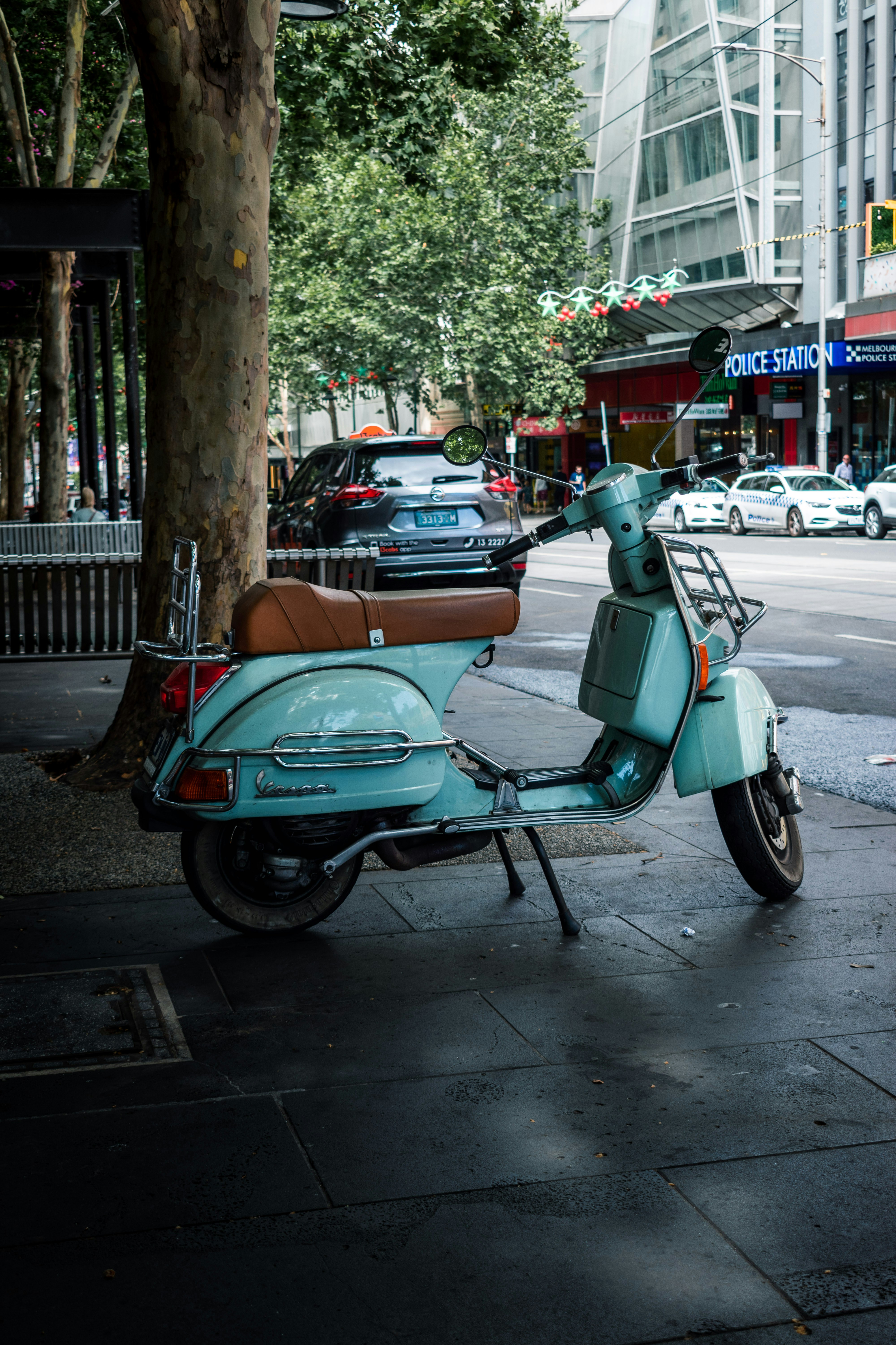 A vintage turquoise scooter parked on a city sidewalk, surrounded by trees and modern architecture. The scene captures a blend of nostalgia and contemporary urban life.