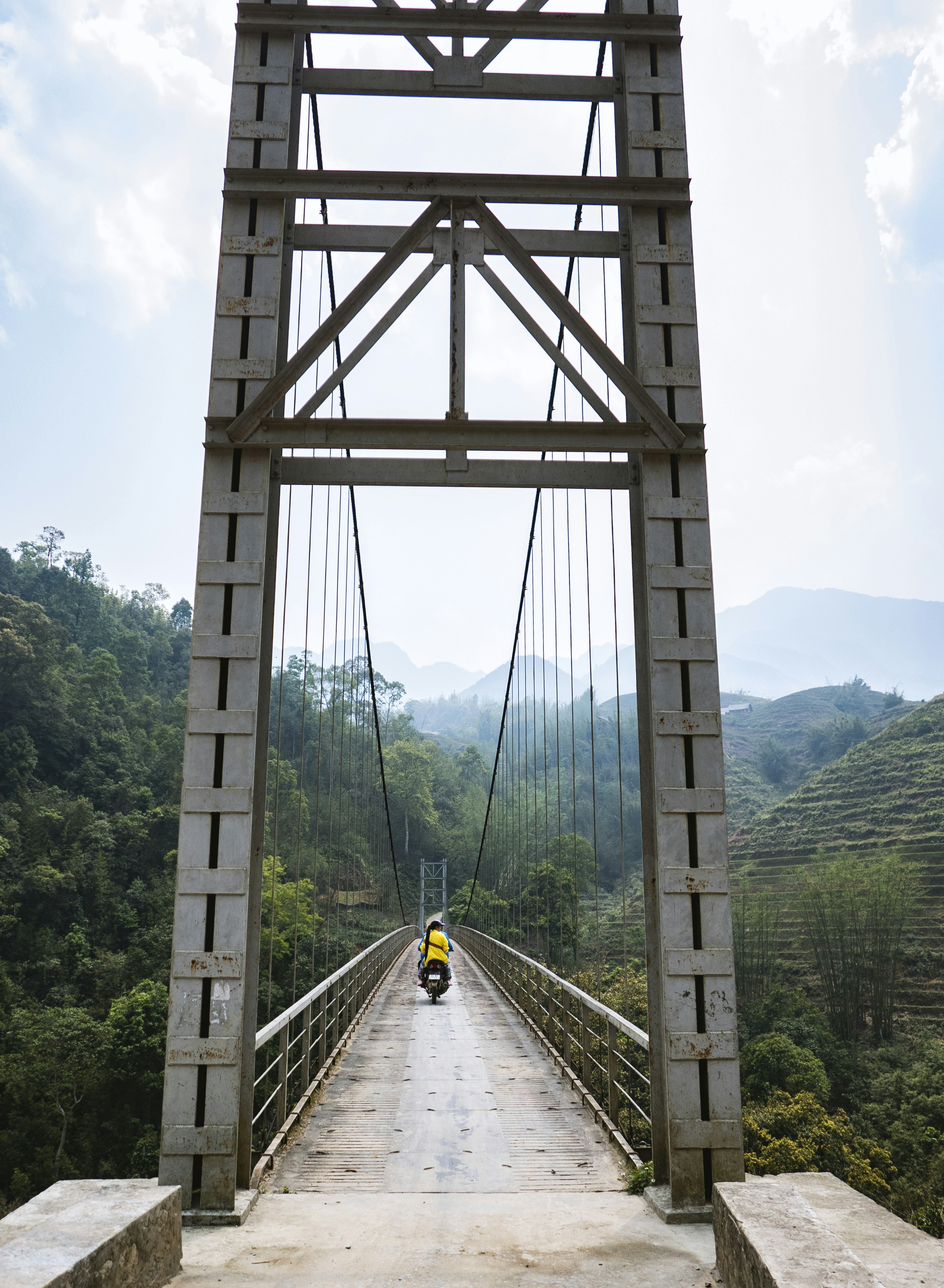 A steel suspension bridge extends toward a lone rider in a bright yellow jacket riding a scooter, crossing a misty valley framed by terraced hills.