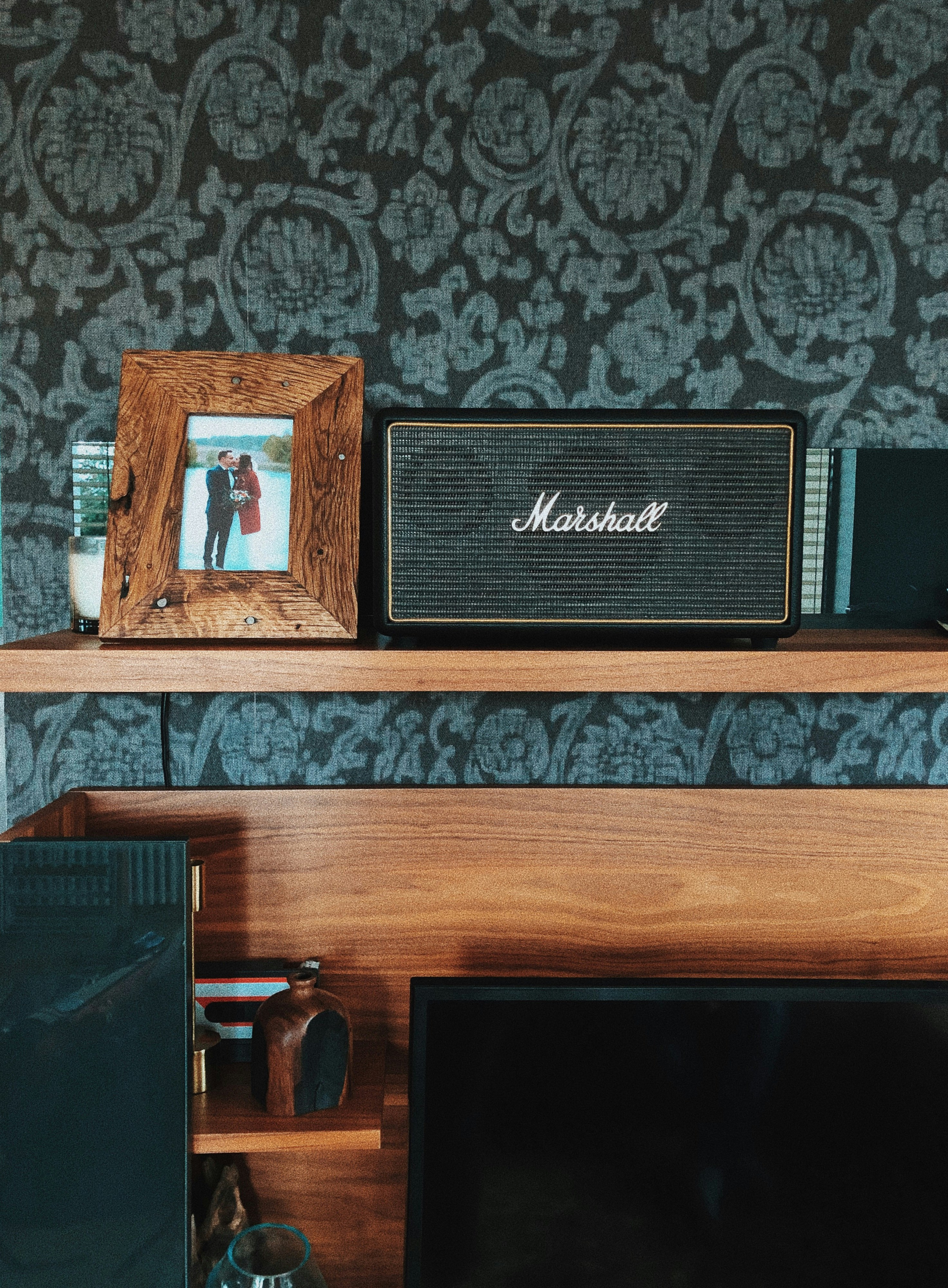 A wooden-framed photo of a couple stands beside a Marshall speaker on a stylish shelf, against a textured floral wallpaper backdrop.
