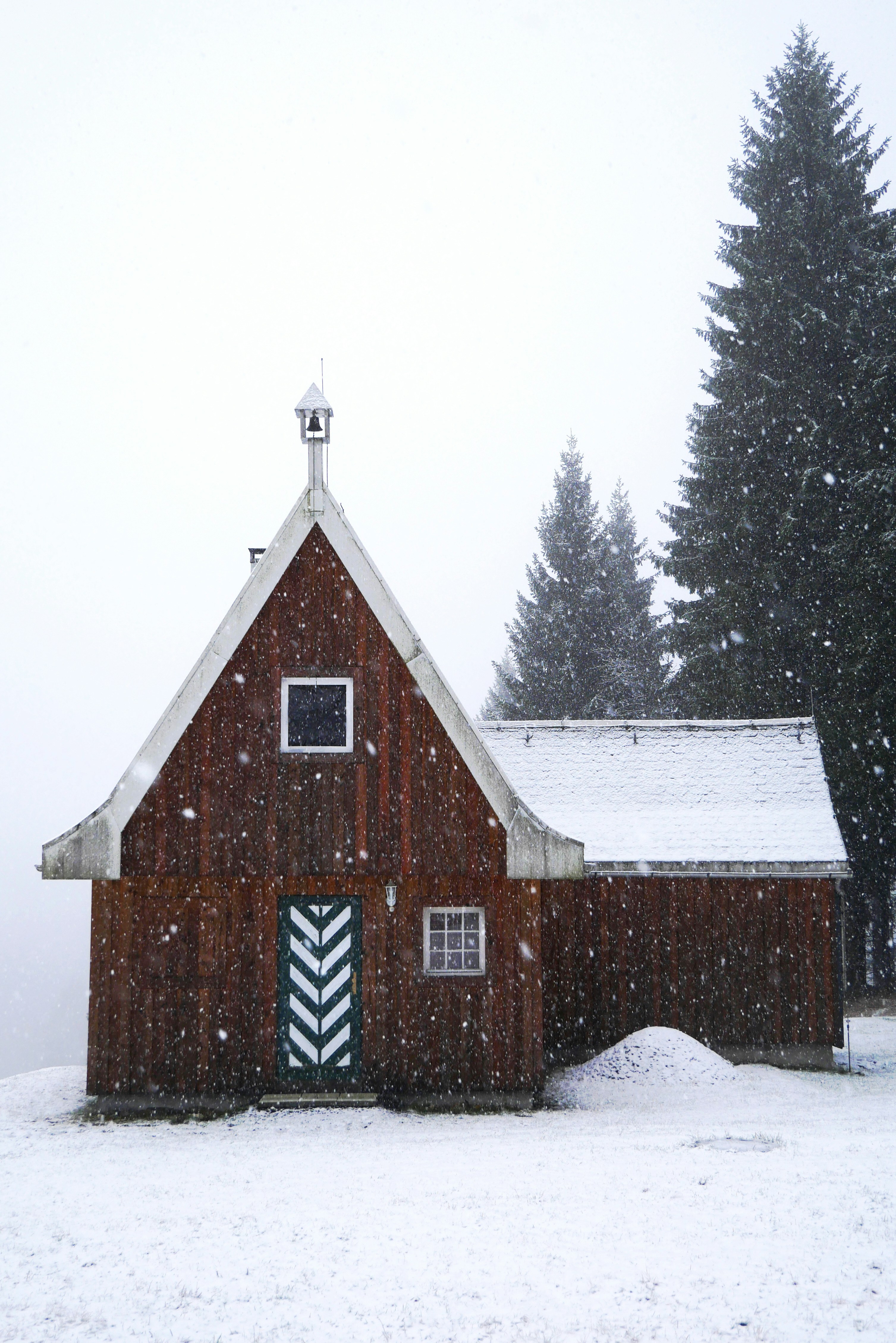 A small red wooden chapel with a white-trimmed steep roof and a green-and-white chevron door stands in a snowy clearing. Snow falls softly as tall evergreen trees rise behind the building.