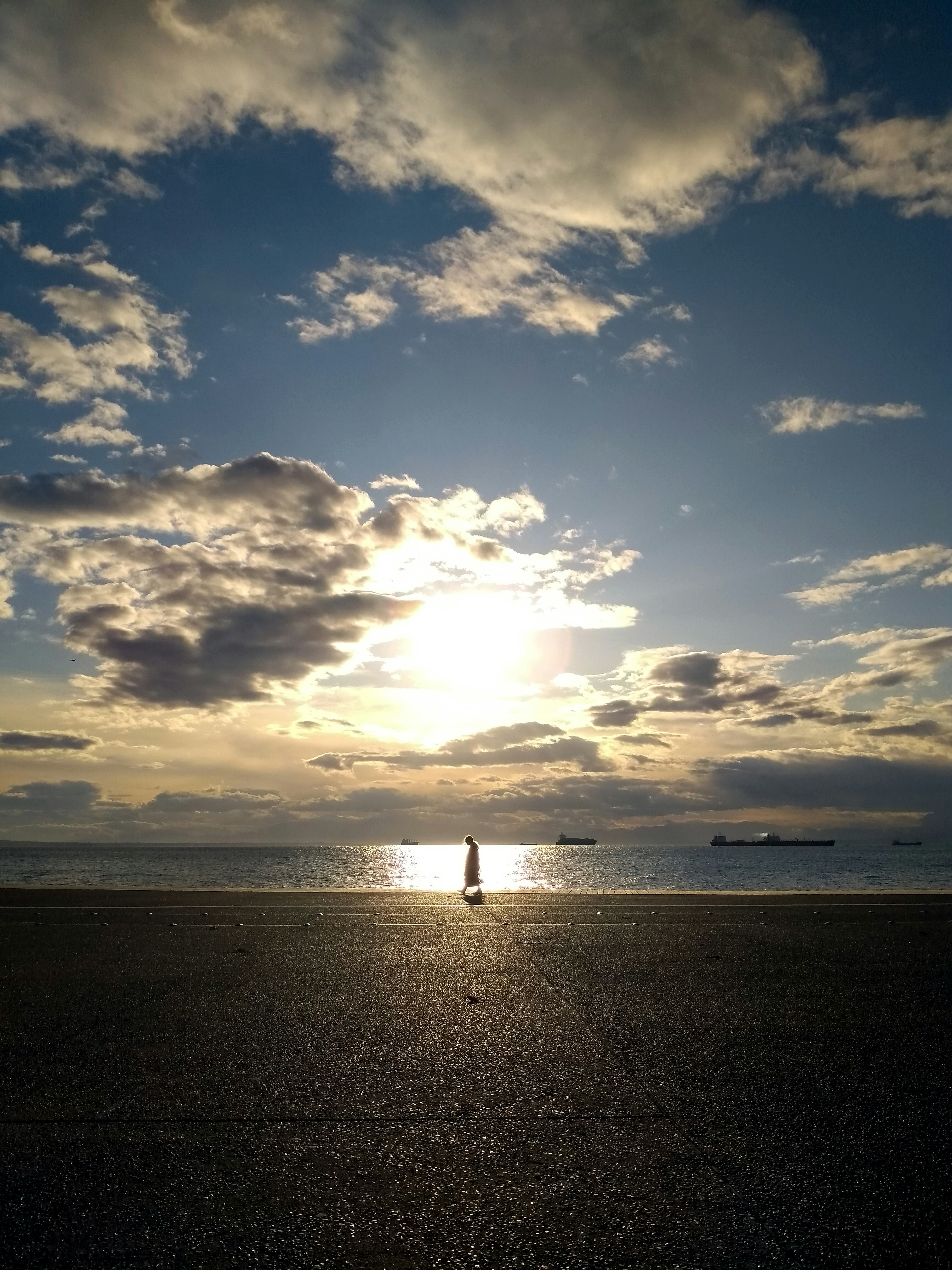 Photograph of a lone figure strolling along a sunlit beach with ships on the horizon. The dramatic sky and shimmering water frame a quiet, contemplative moment.