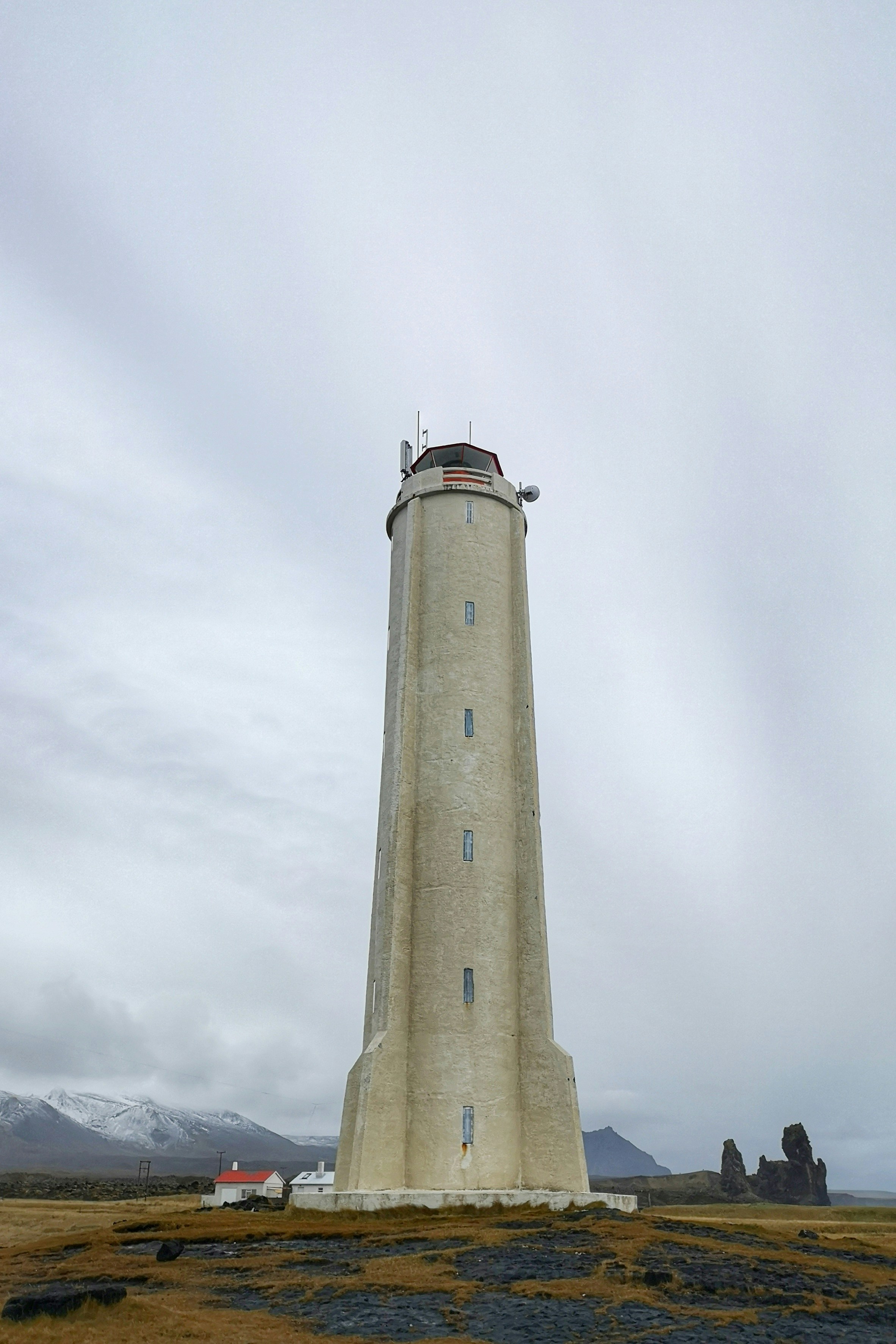 Tall lighthouse standing against a backdrop of gray skies and distant mountains, with a small building at its base.