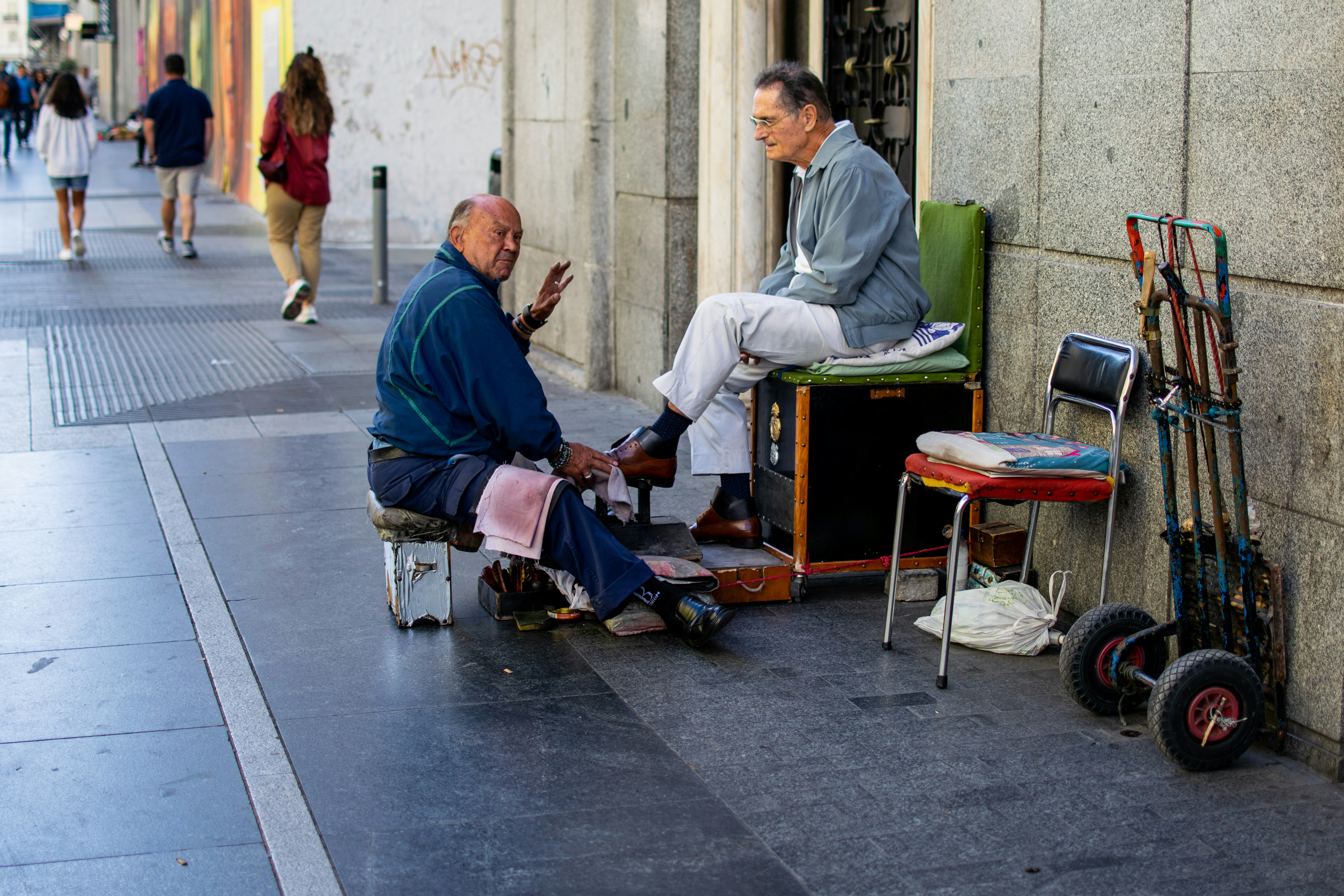 Two men engaged in conversation on a bustling city sidewalk, surrounded by everyday urban elements. The scene captures a moment of connection amidst the city's rhythm.