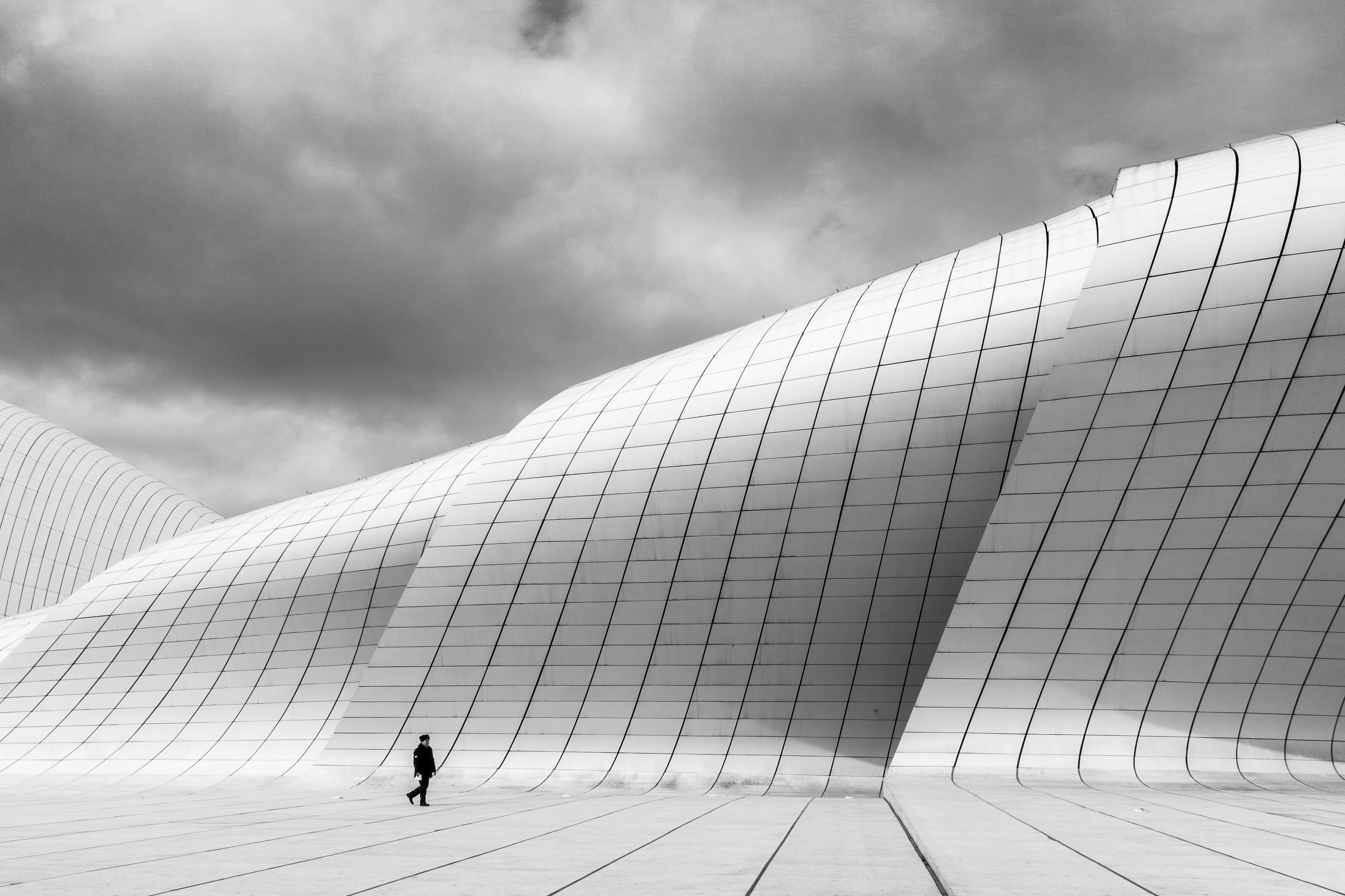 Person walking beside a curving, grid-patterned building under dramatic clouds.