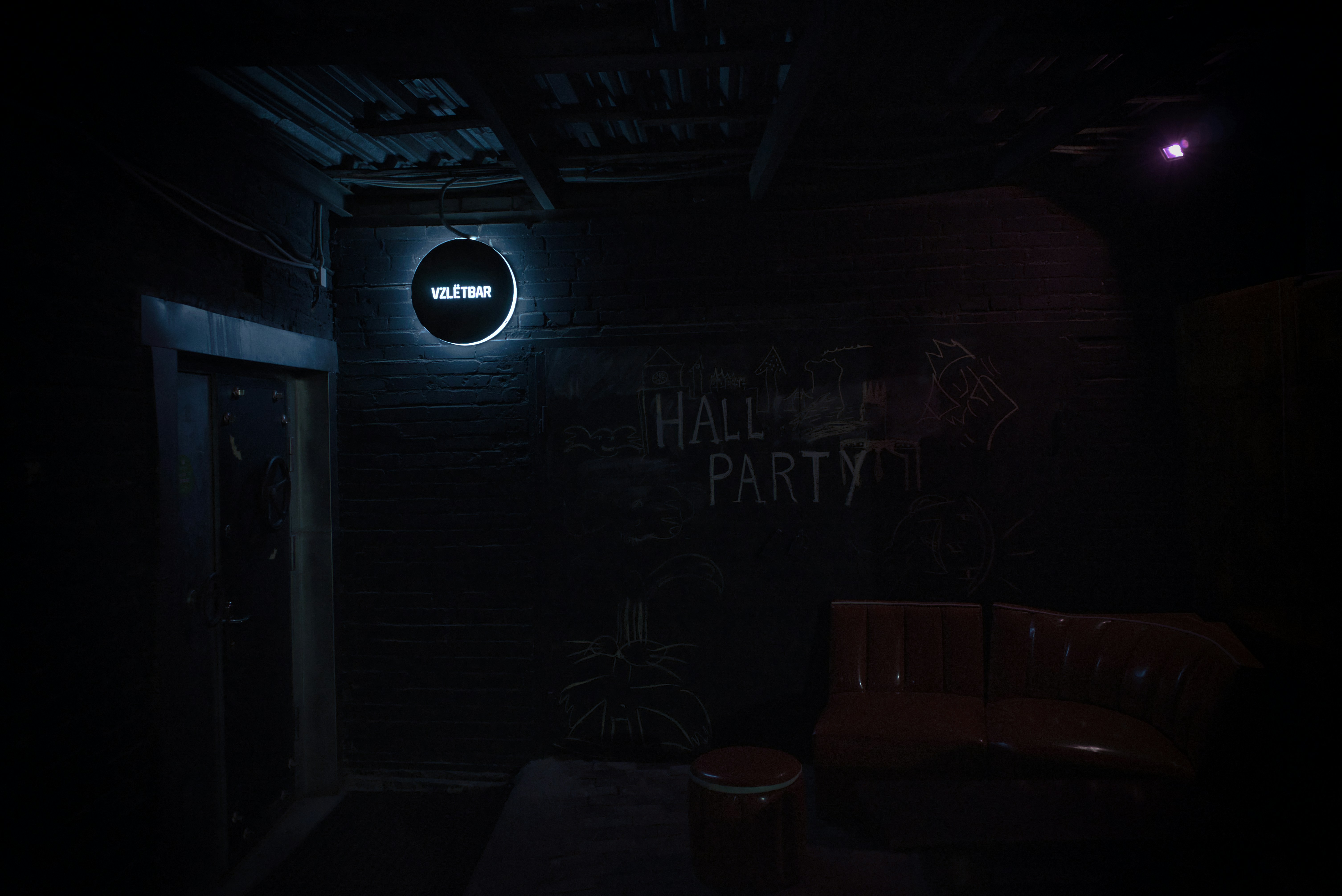 A dimly lit room featuring a glowing neon sign that reads 'VOLTAM', with a vintage couch and chalkboard art in the background.