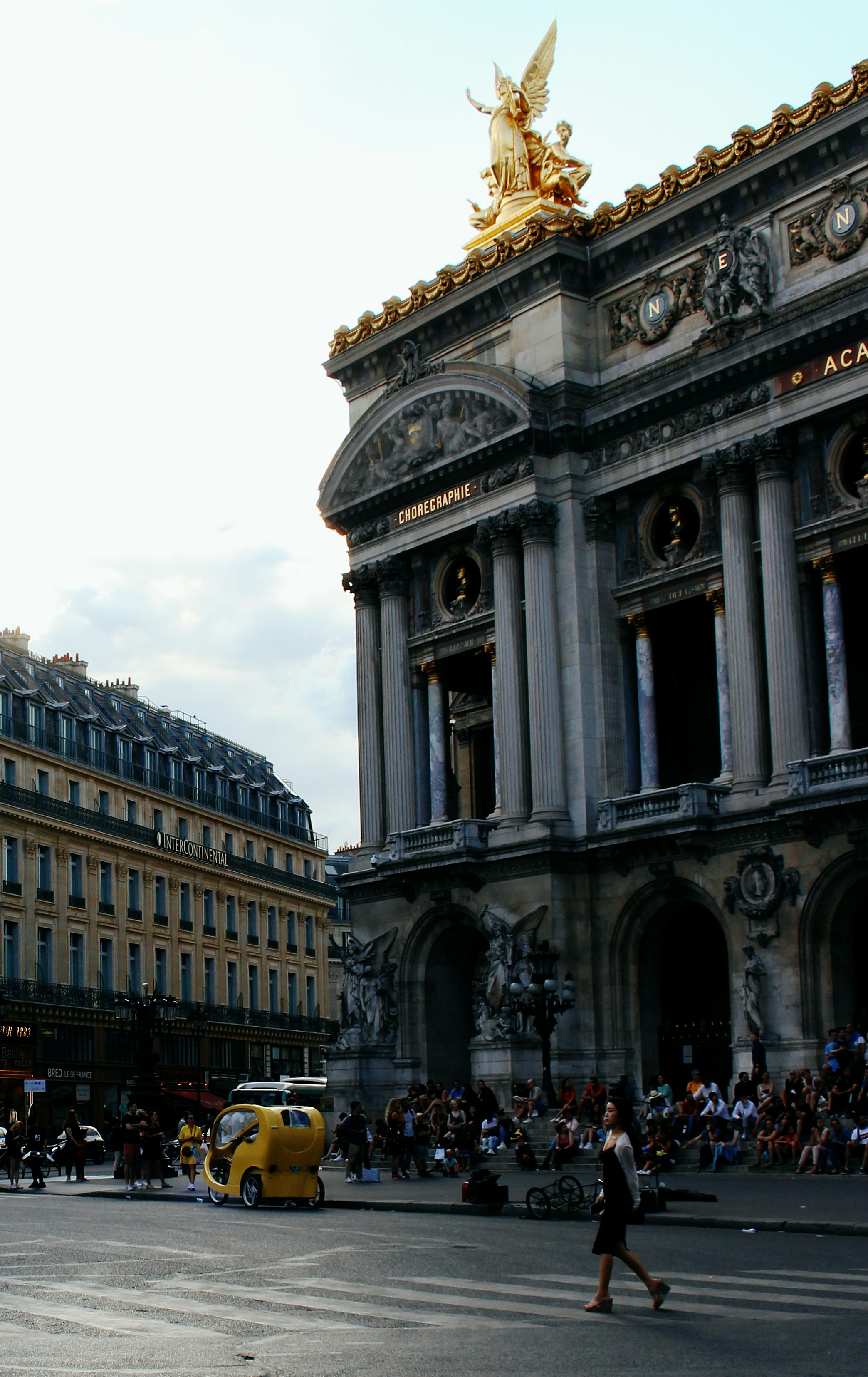 The ornate façade of the Palais Garnier, adorned with a golden statue, stands majestically against a cloudy sky, capturing the essence of Parisian architecture. A vibrant yellow vehicle adds a modern touch to the historic scene.