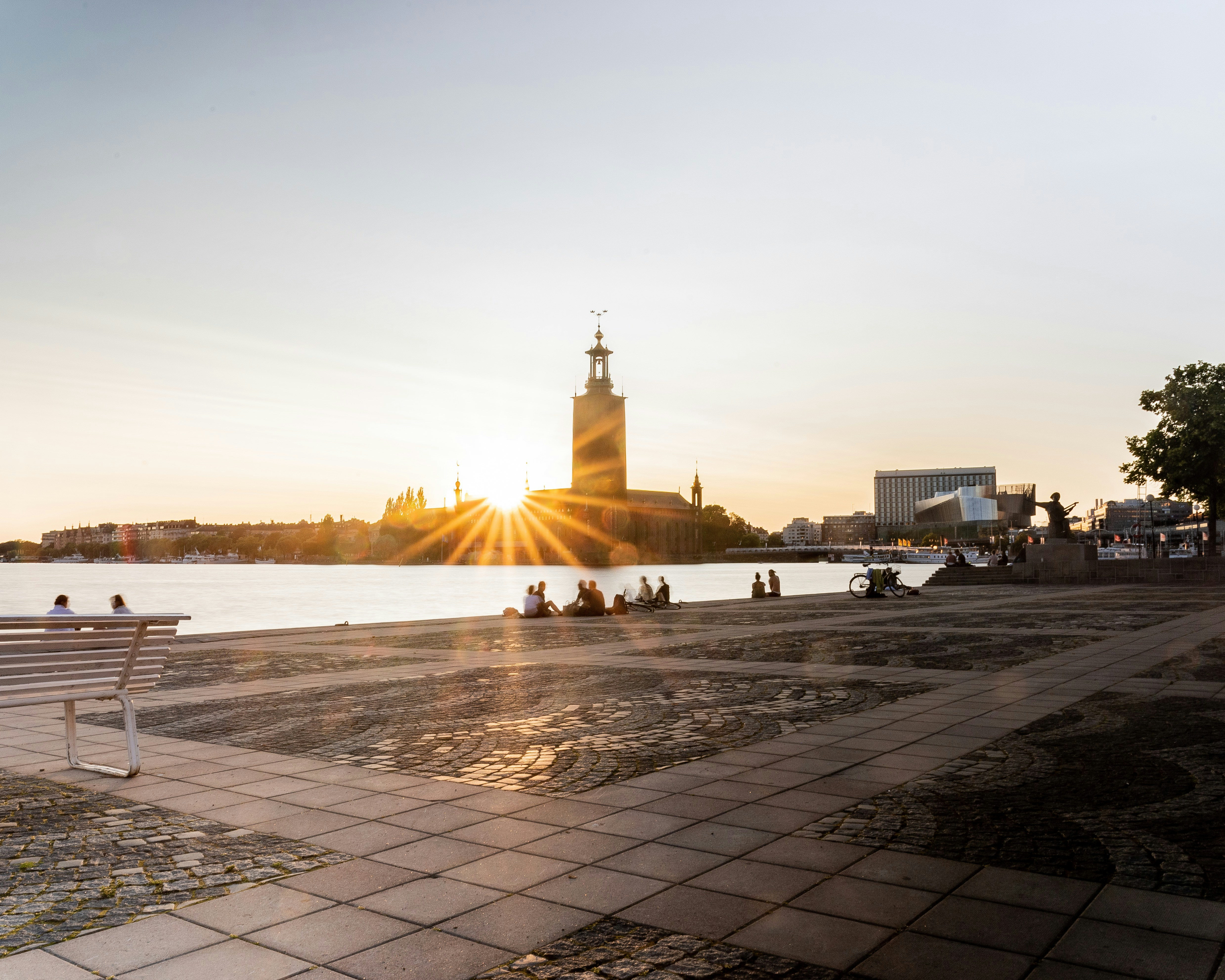 Silhouetted figures seated by a waterfront, gazing at the sun setting behind a cityscape with a prominent tower.