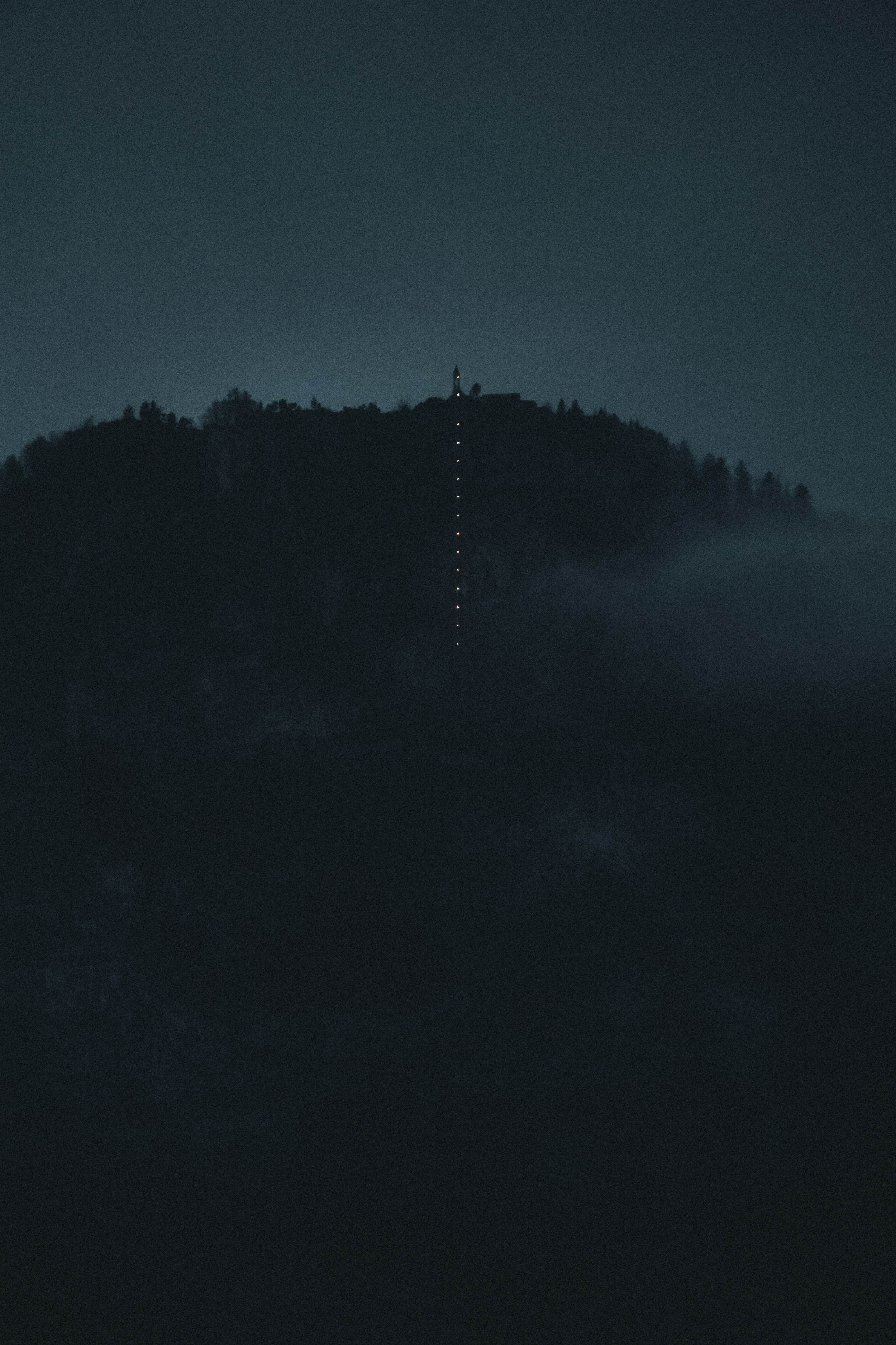 A solitary figure stands atop a mountain, illuminated by a vertical line of lights against a dark, moody sky. The scene captures the essence of solitude and adventure.